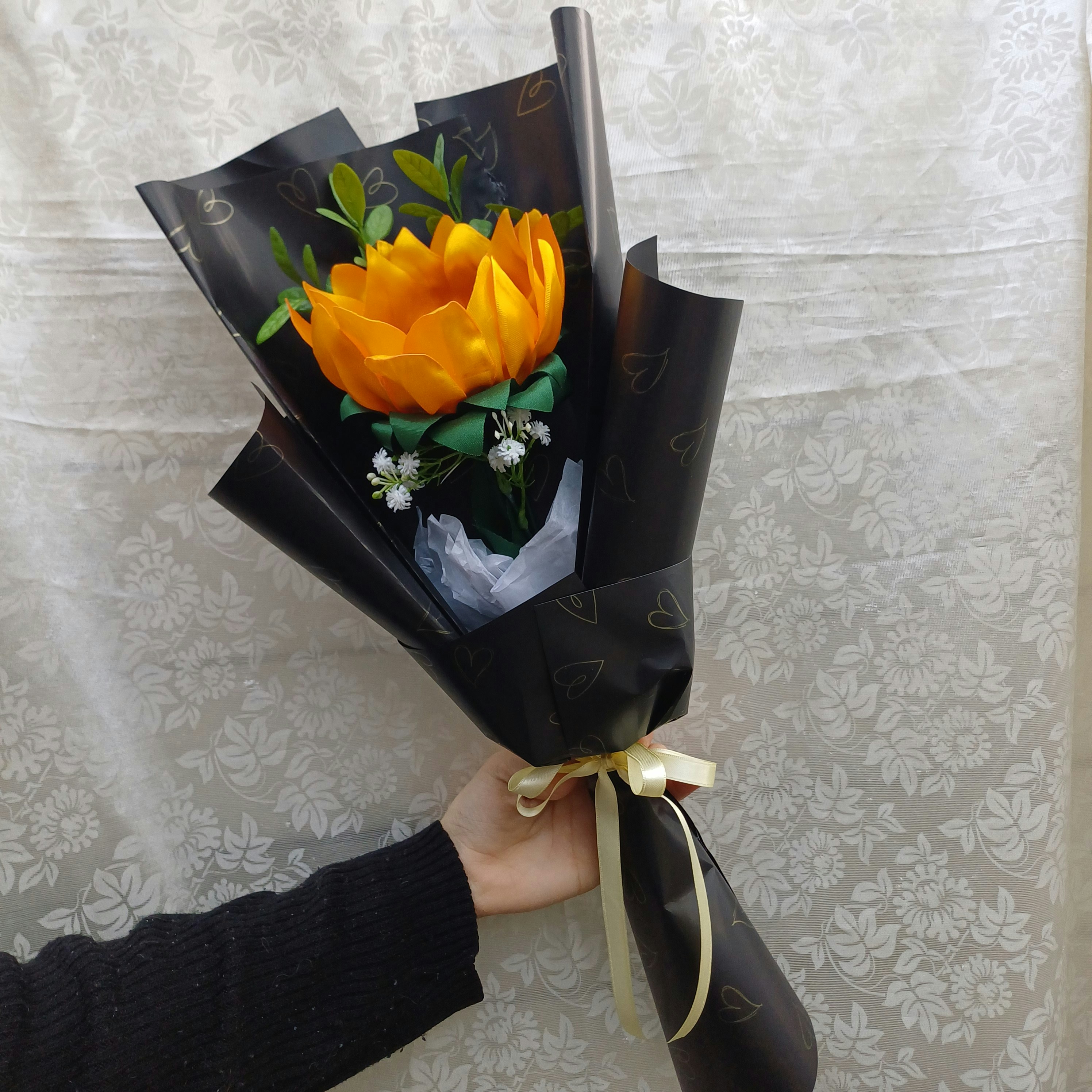 Bright yellow bouquet wrapped in black paper held by a gloved hand against a pale patterned backdrop. Floral arrangement appears as the focal point with natural lighting and a soft texture surrounding it.