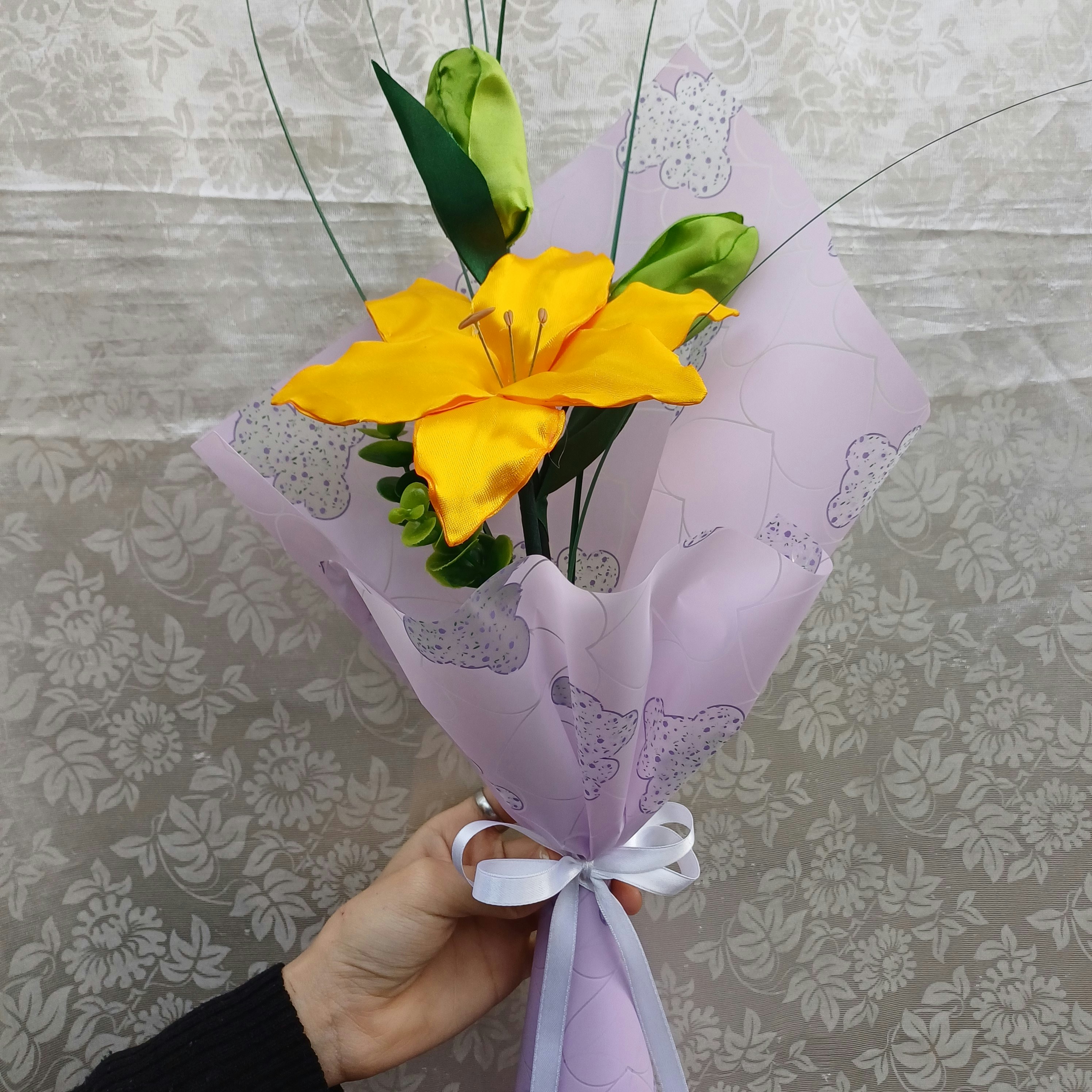 A hand holding a yellow fabric lily bouquet wrapped in patterned lavender paper