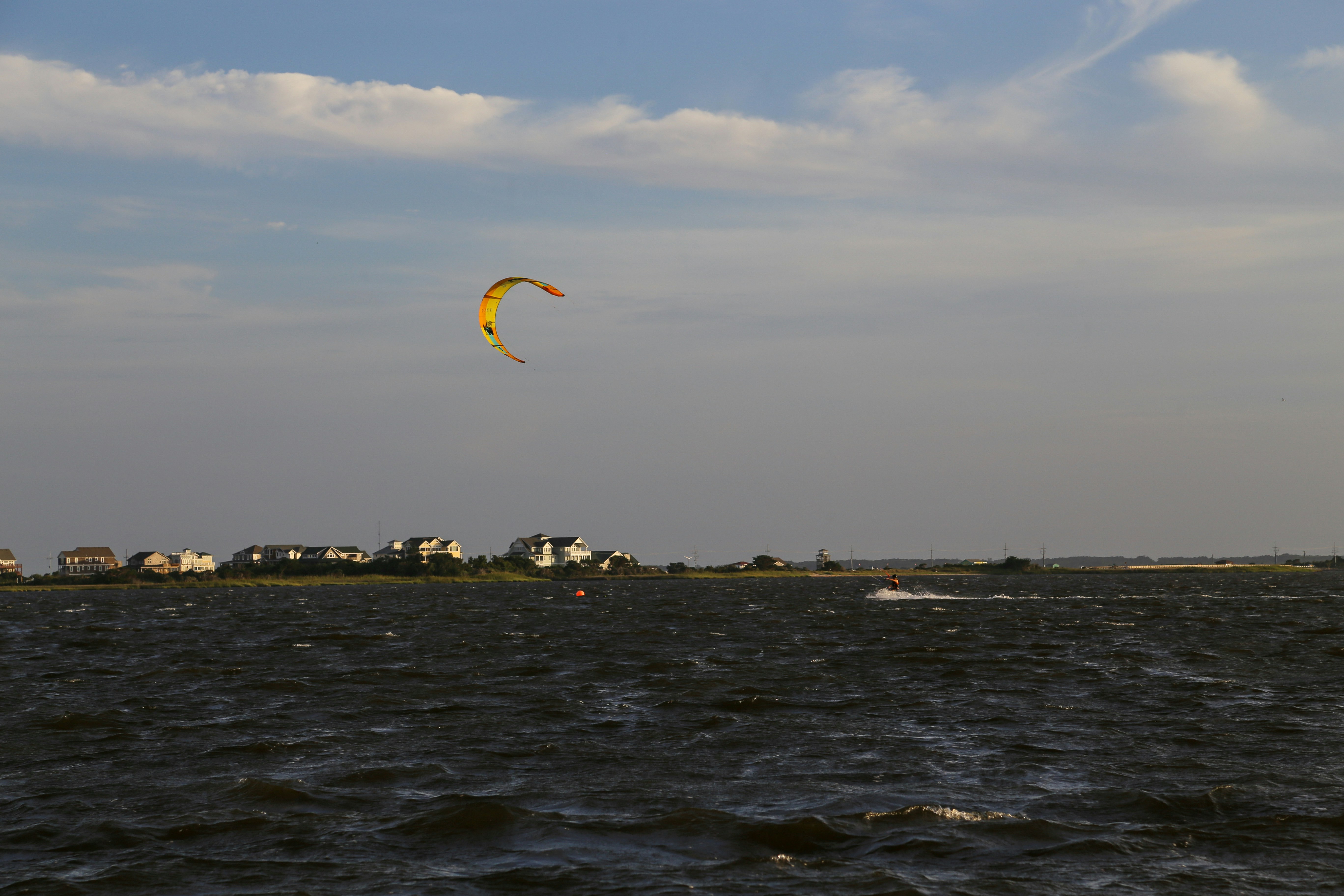 Kite surfer over choppy water near a distant coastal row of houses, under a blue sky with scattered clouds.