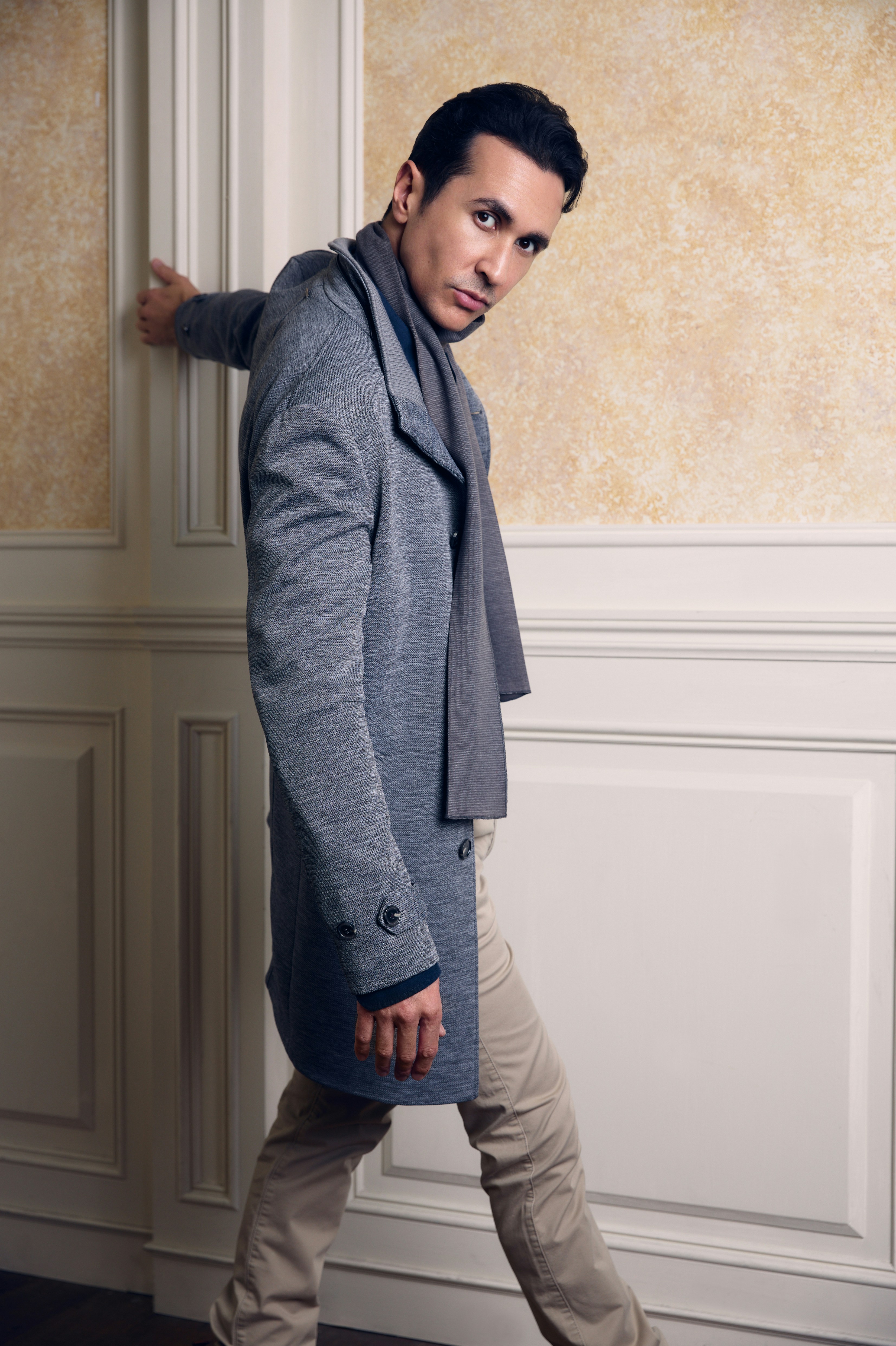 Slim man in a light gray coat leans against a doorway in a softly lit hallway with cream paneling.