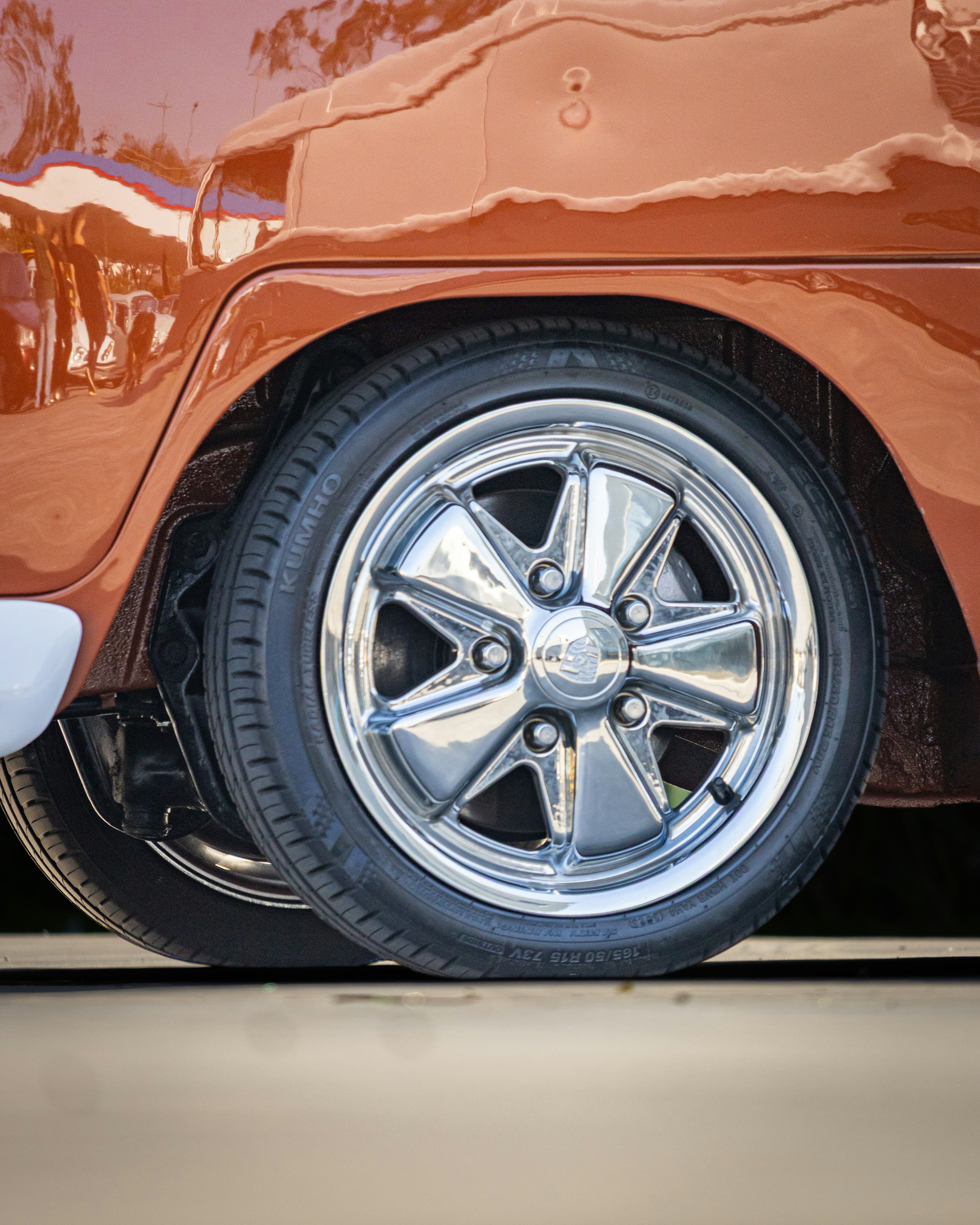 A close-up of a chrome car wheel set into a brown-painted fender, showcasing polished rims and tire tread with reflections on the surrounding surface.