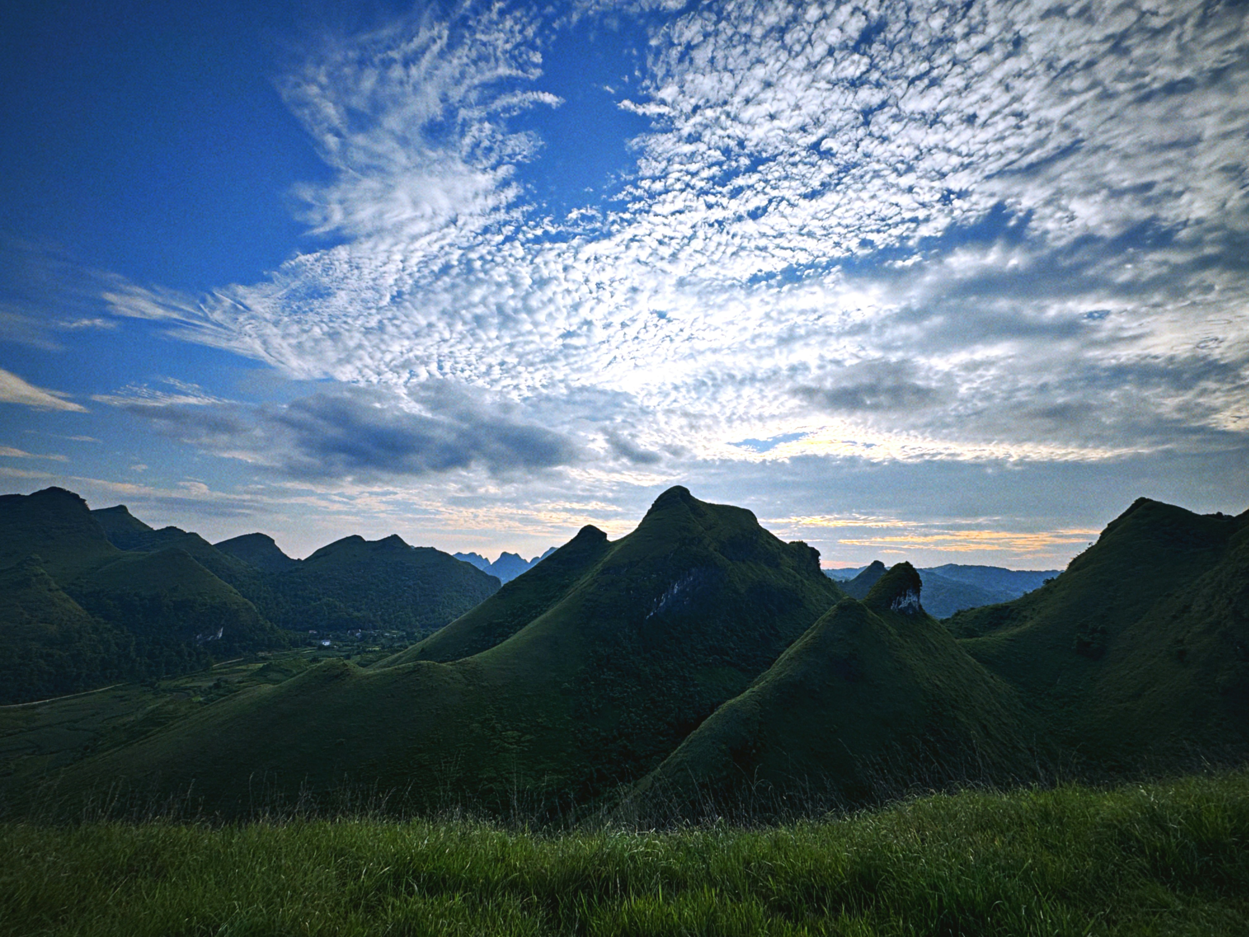 Undulating green hills roll toward a distant crest under a textured sky; the scene captures expansive landscape with a broad, relaxed composition.