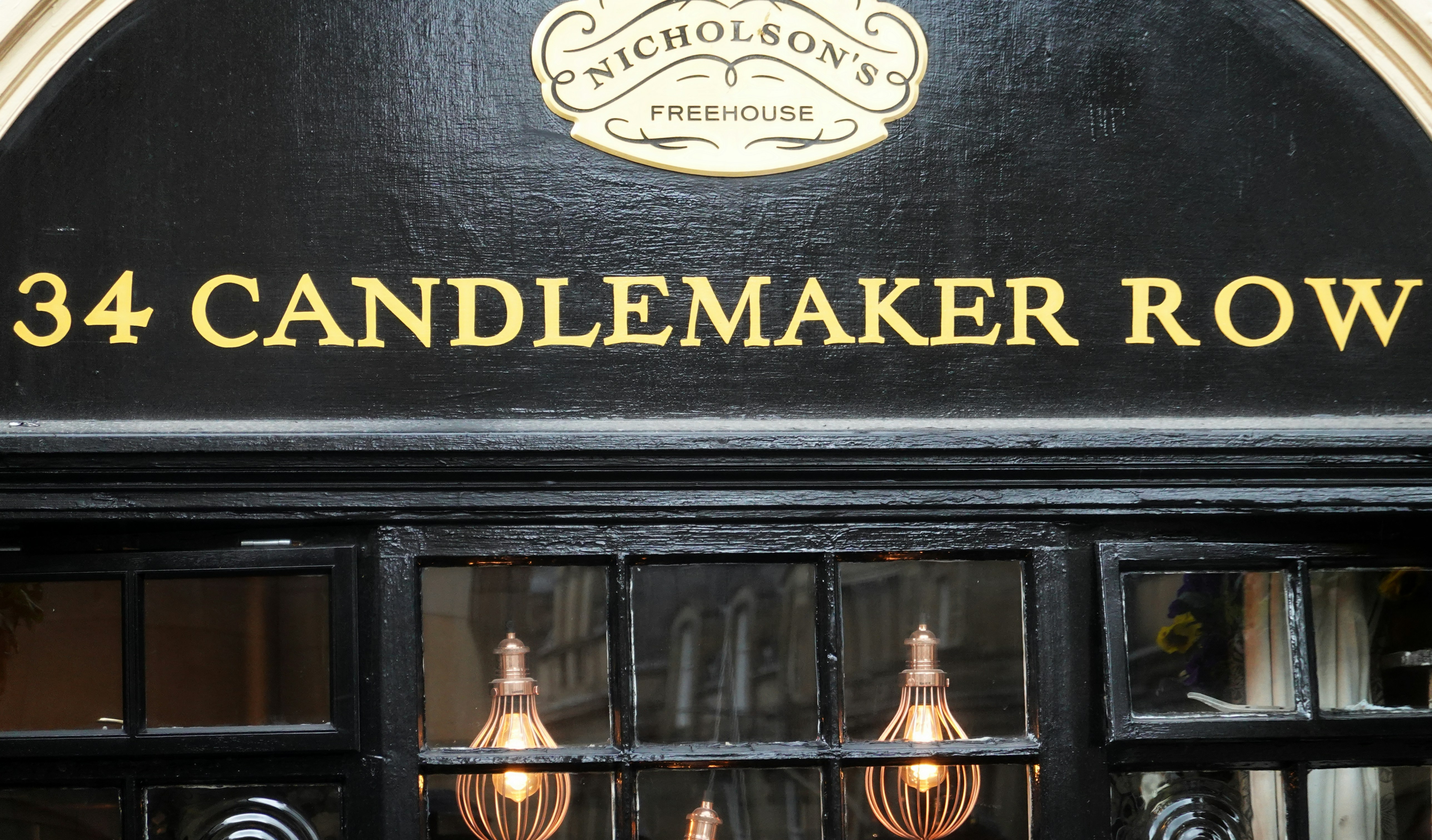 Dark storefront with large glass panes and warm filament bulbs visible through the window; a vintage sign above reads ‘34 Candlemaker Row’.