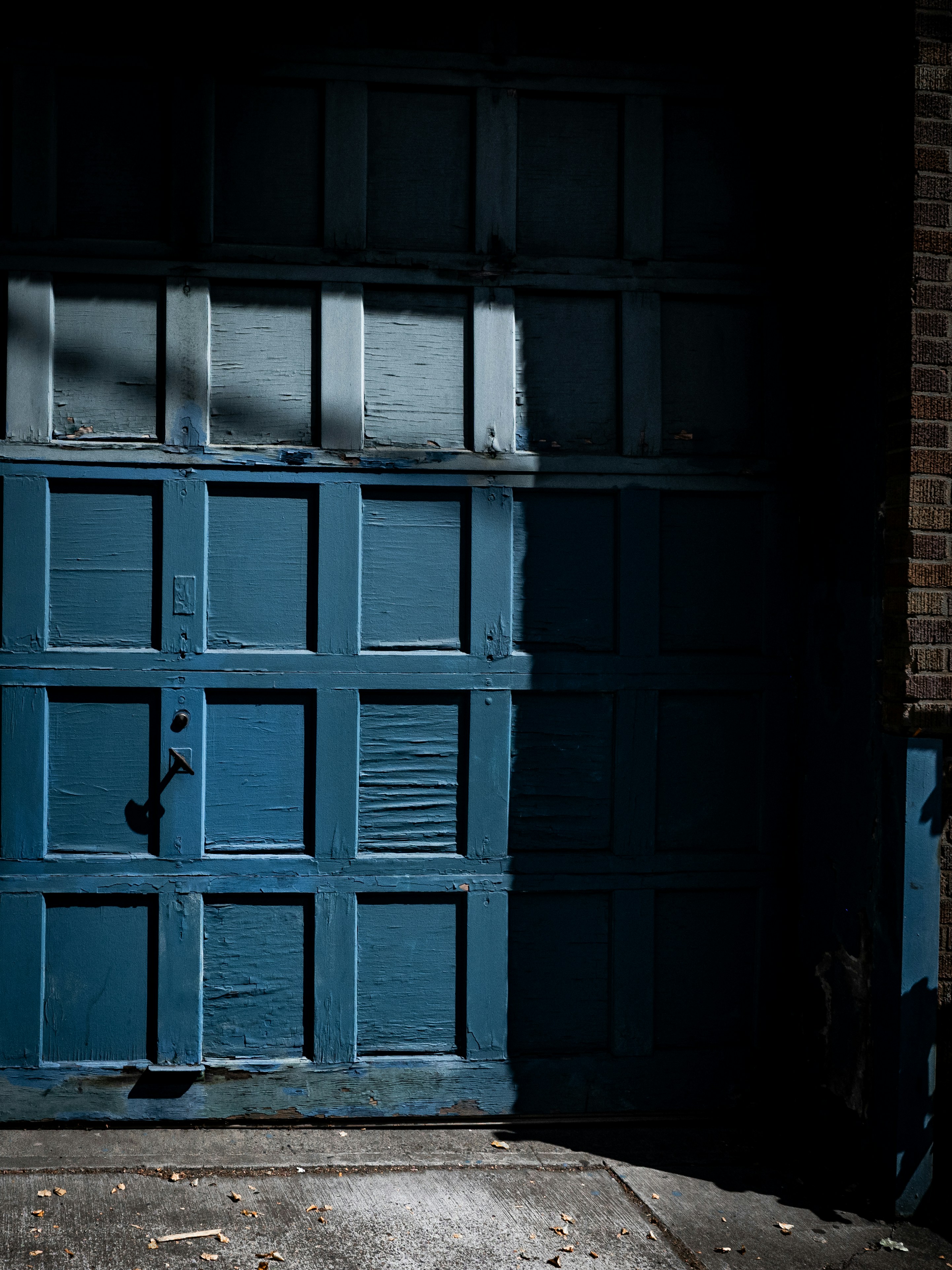 Blue wooden garage door with square panels, bathed in low, directional light that reveals texture and wear; a hint of brick wall on the right adds contrast.