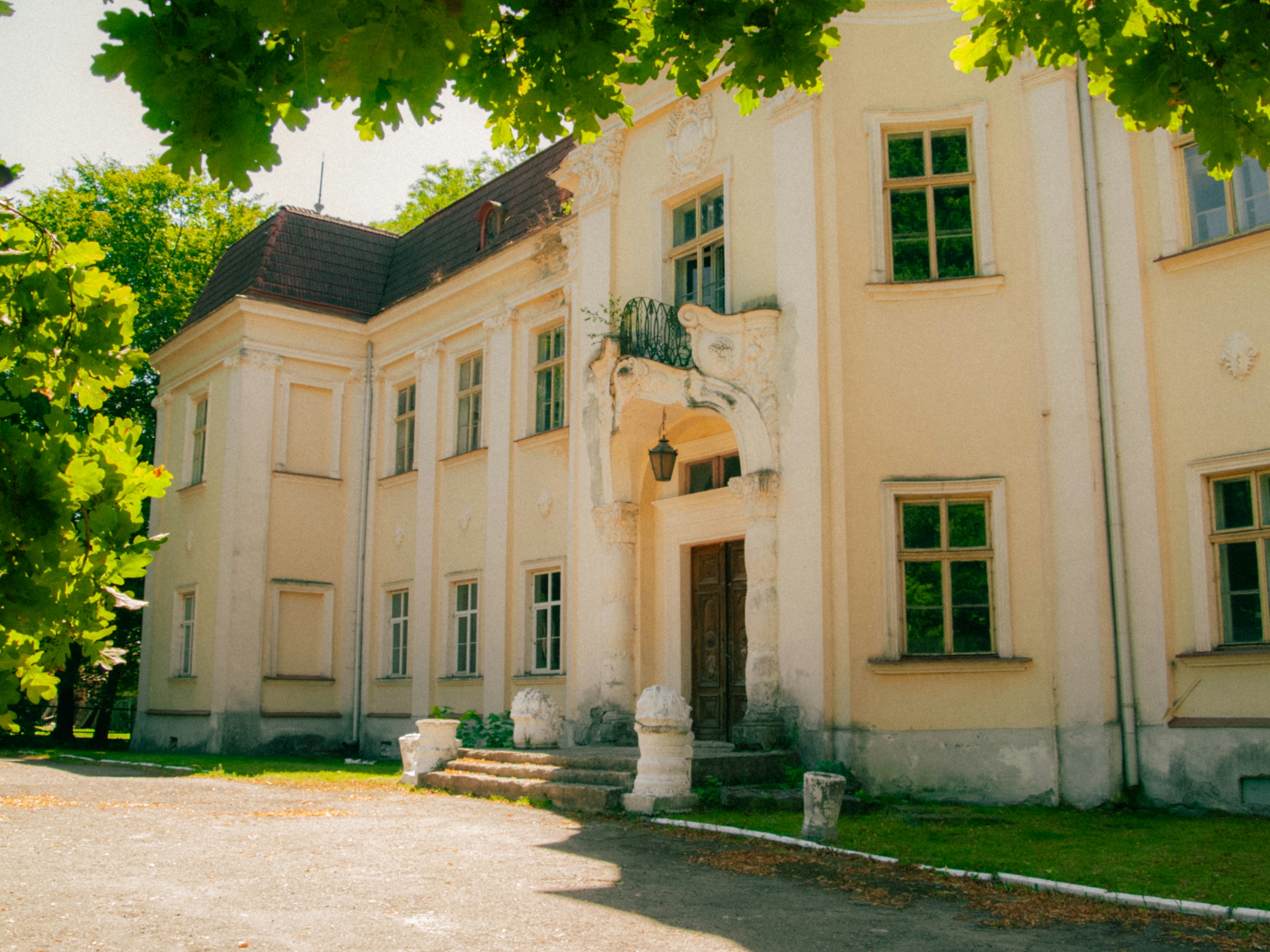 Sunlit historic villa façade with carved doorway and arched entry, framed by green trees; a tranquil, daylight street scene emphasizing architecture and texture.