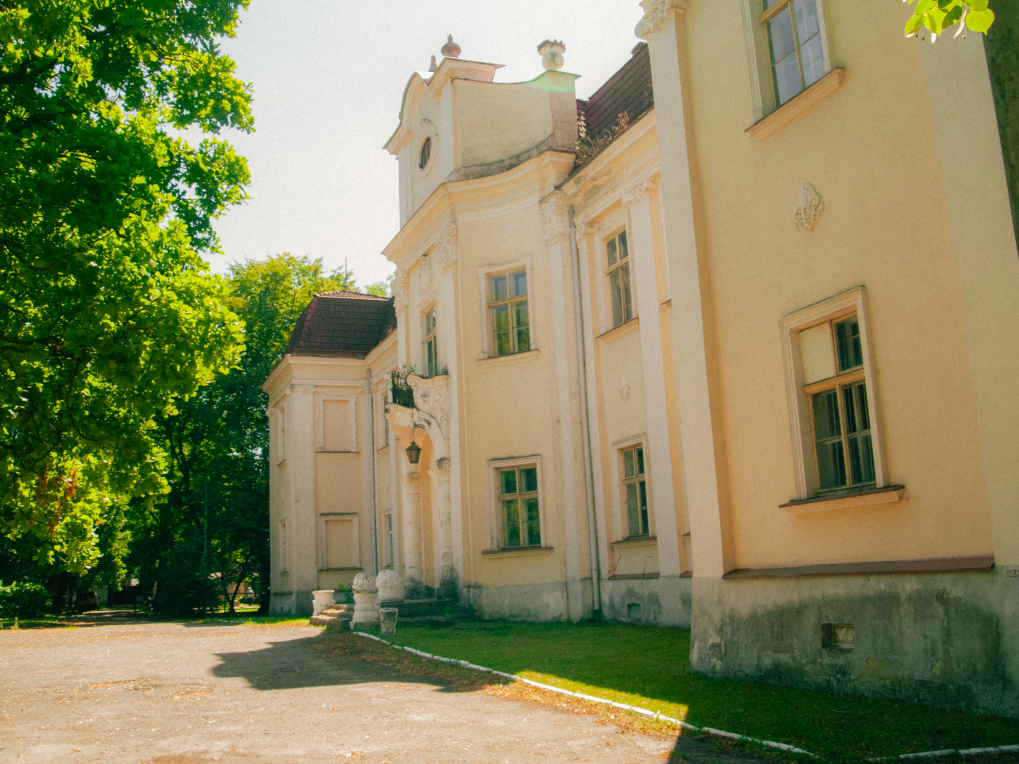 Sunlit pale-yellow villa facade with arched windows and lush green trees beside a paved courtyard. A calm moment showcasing classic architecture and gentle shadows.