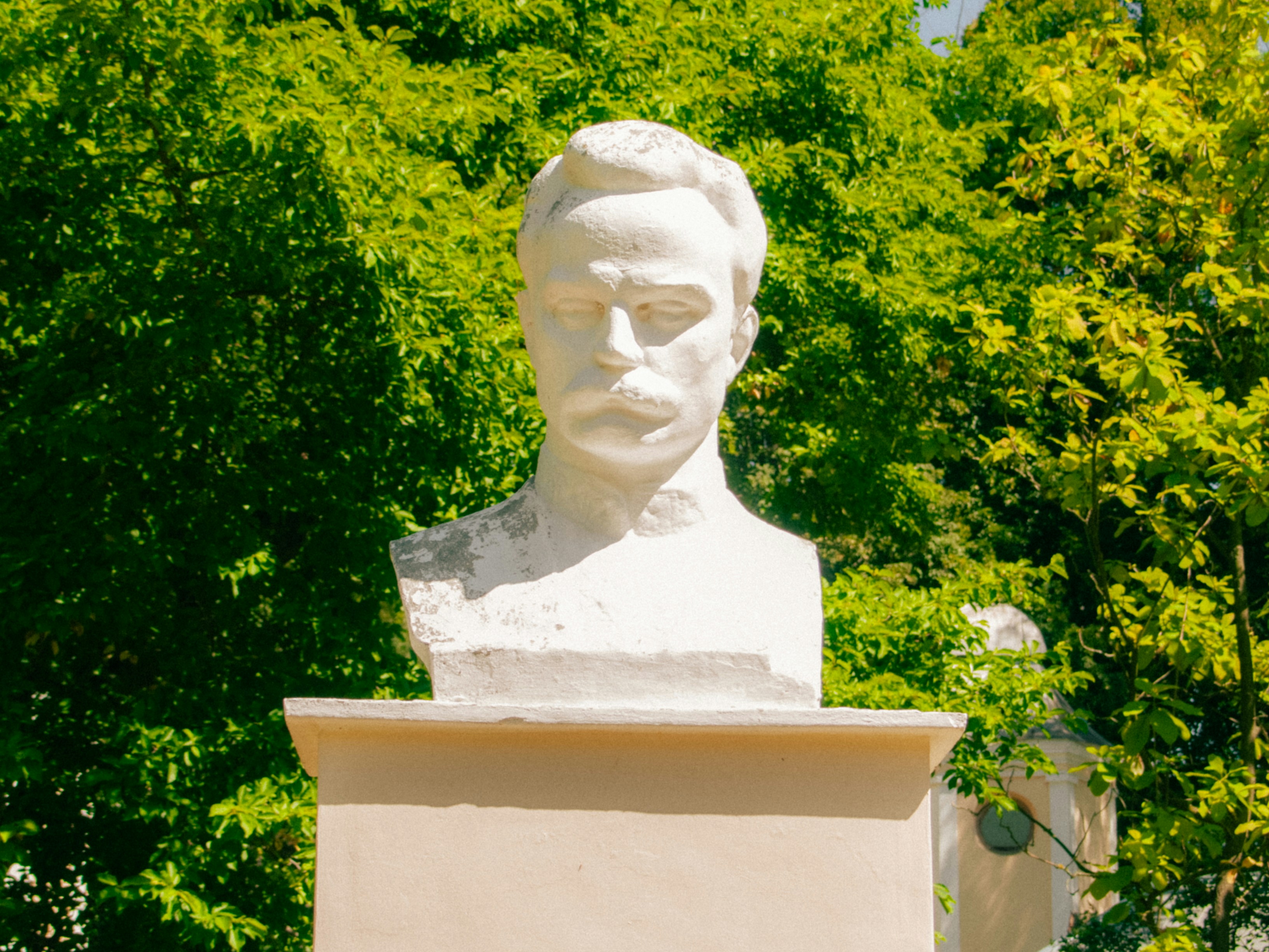 White bust on pedestal framed by vibrant green trees in a sunlit garden.