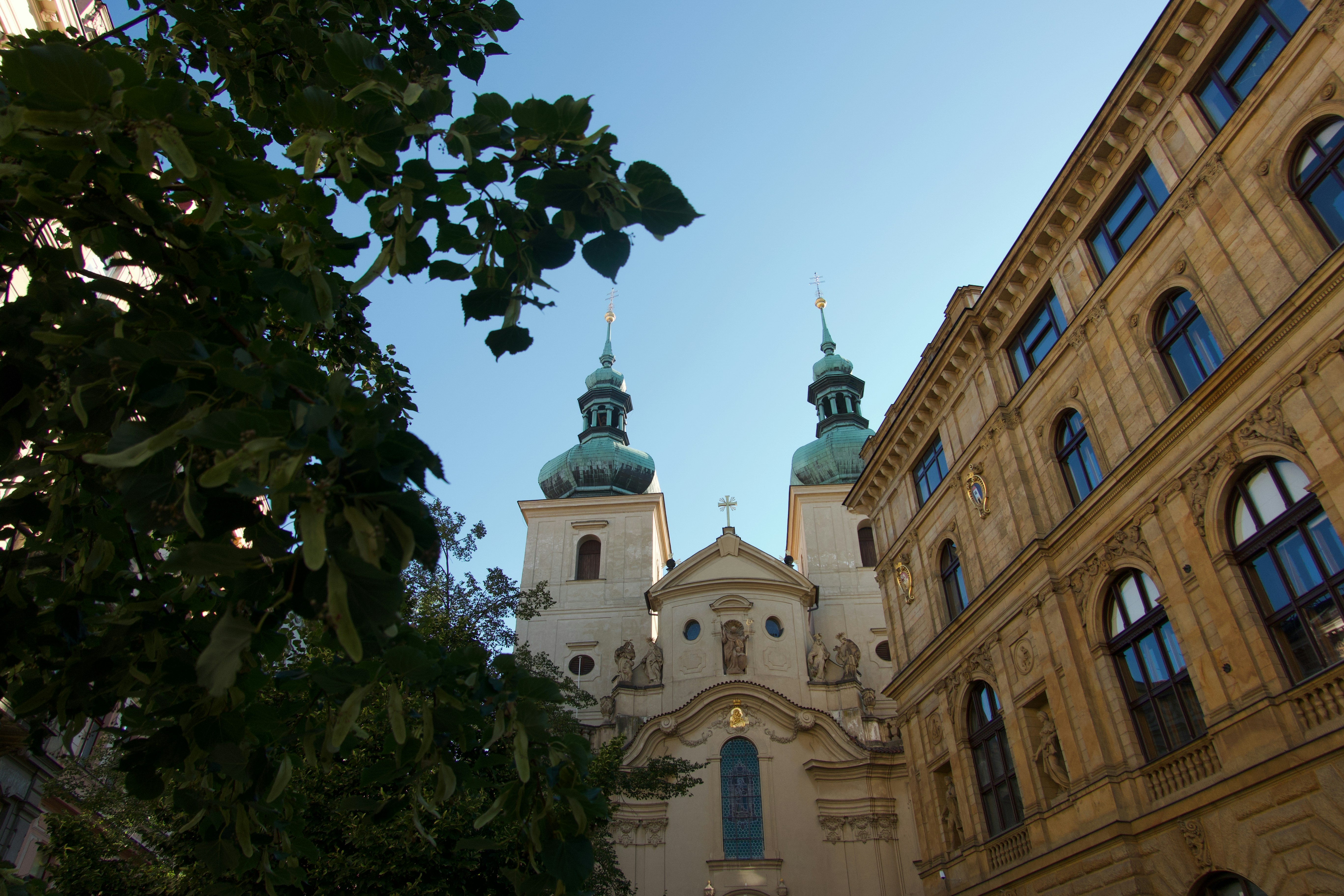 Sunlit church facade rises between tree canopy and a classical brick building, highlighting its twin green domes and ornate central pediment.