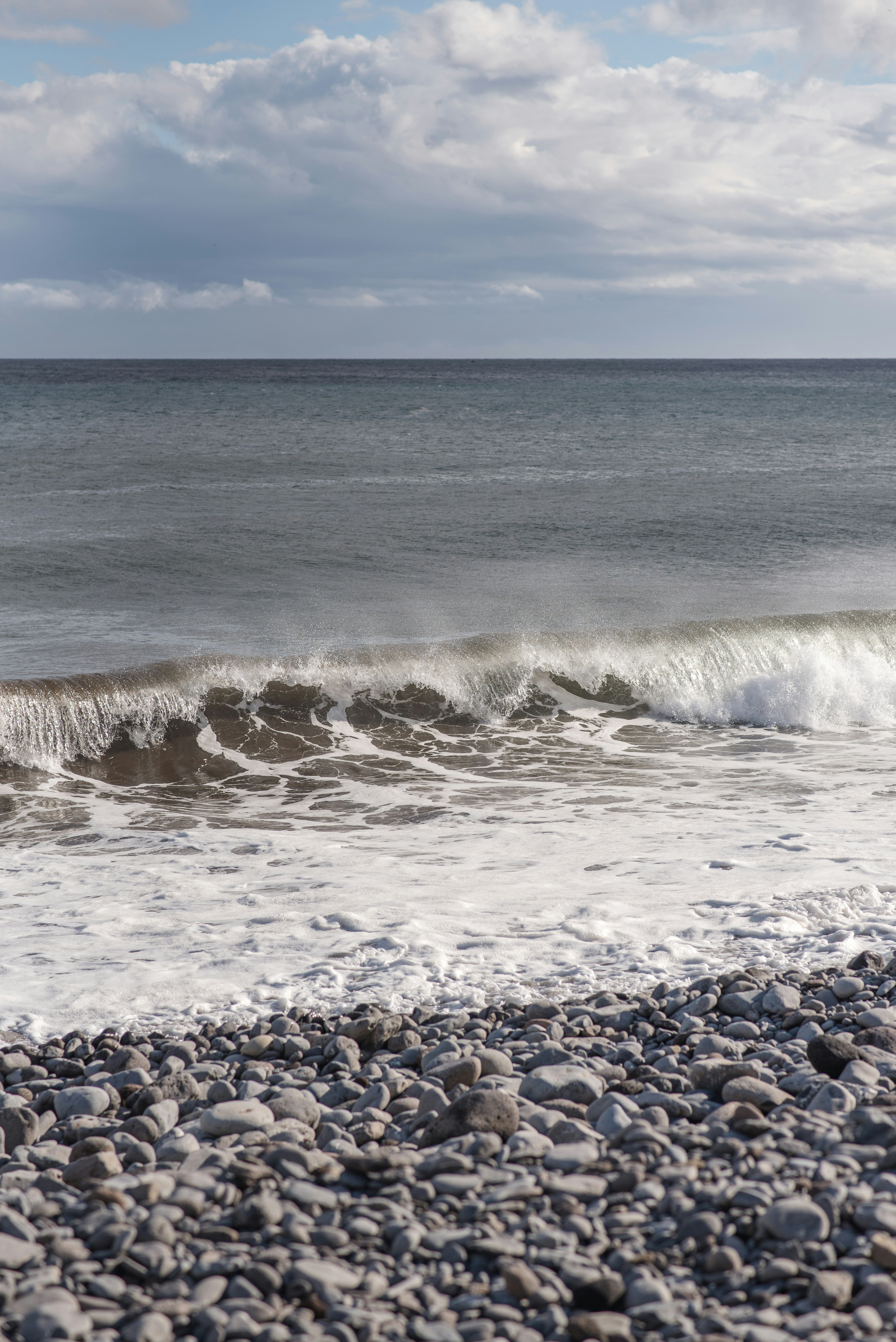 Pebble beach along a calm sea with rolling waves and a cloudy sky, emphasizing texture of the stones and the horizon line.