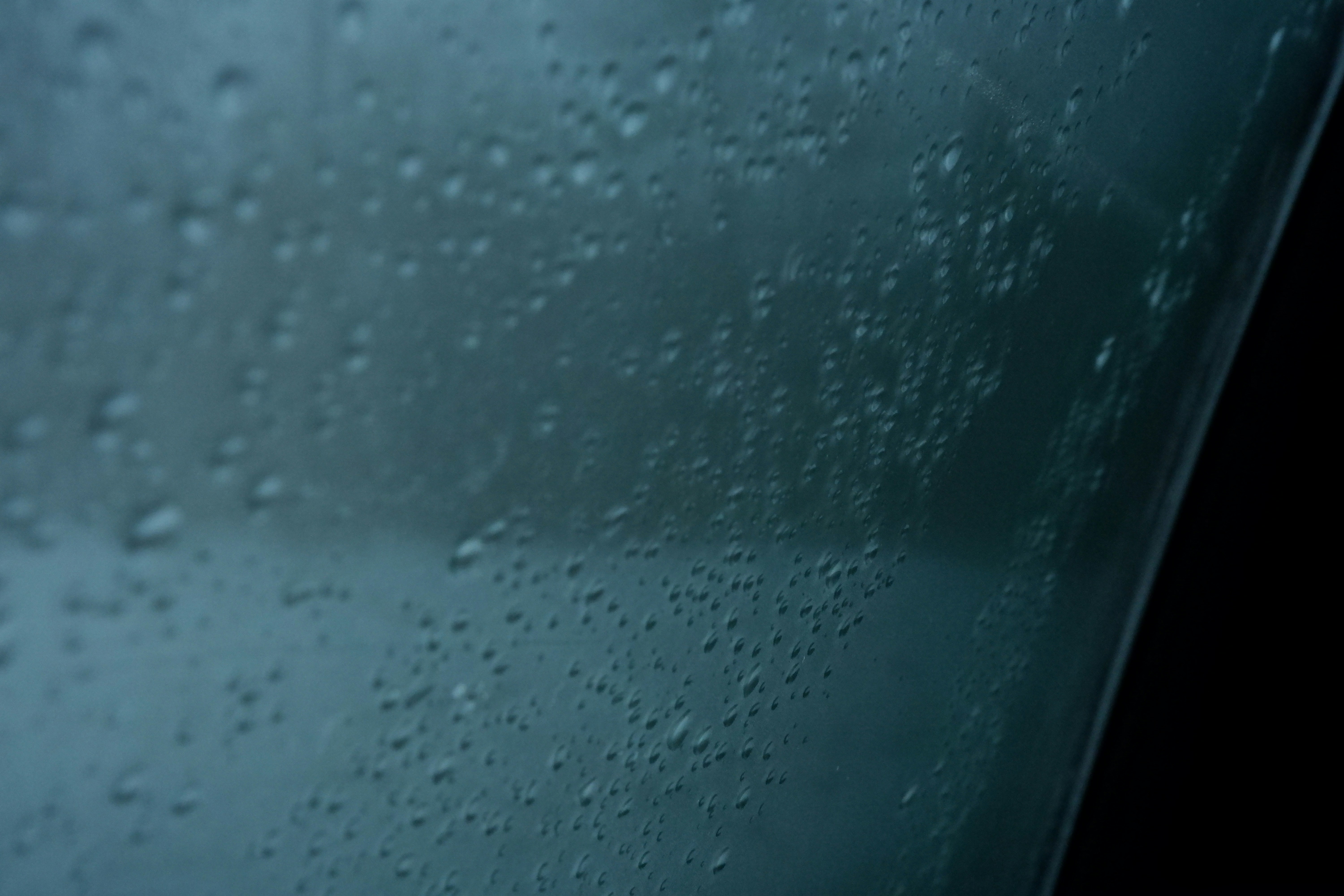 Close-up of a rain-splashed glass pane with water droplets diffusing light, set against a dark edge; minimalist abstract texture that hints at a window during rain.