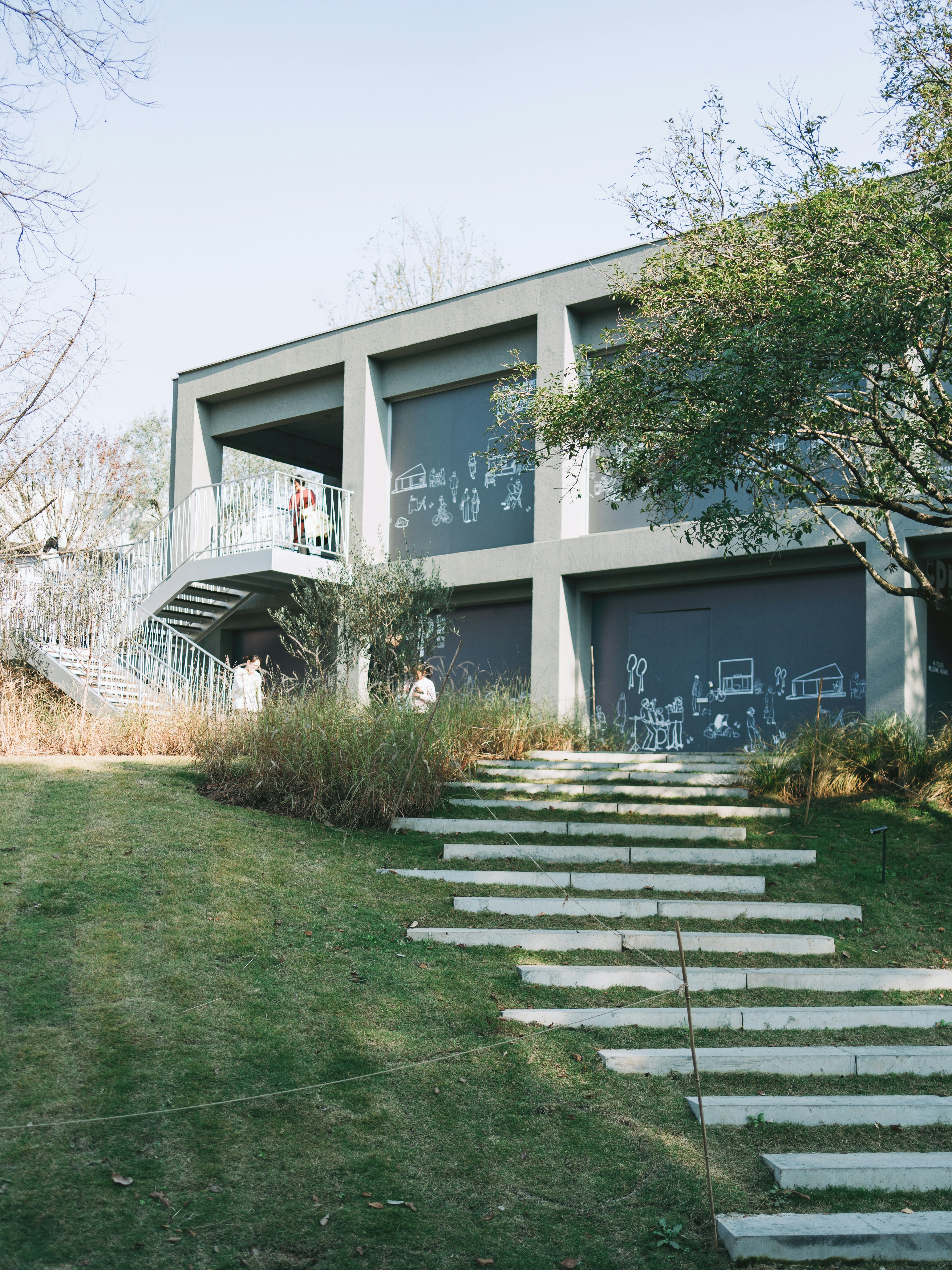 Concrete modern building with chalk drawings on large blue garage doors, stairs leading up, and greenery in the foreground.