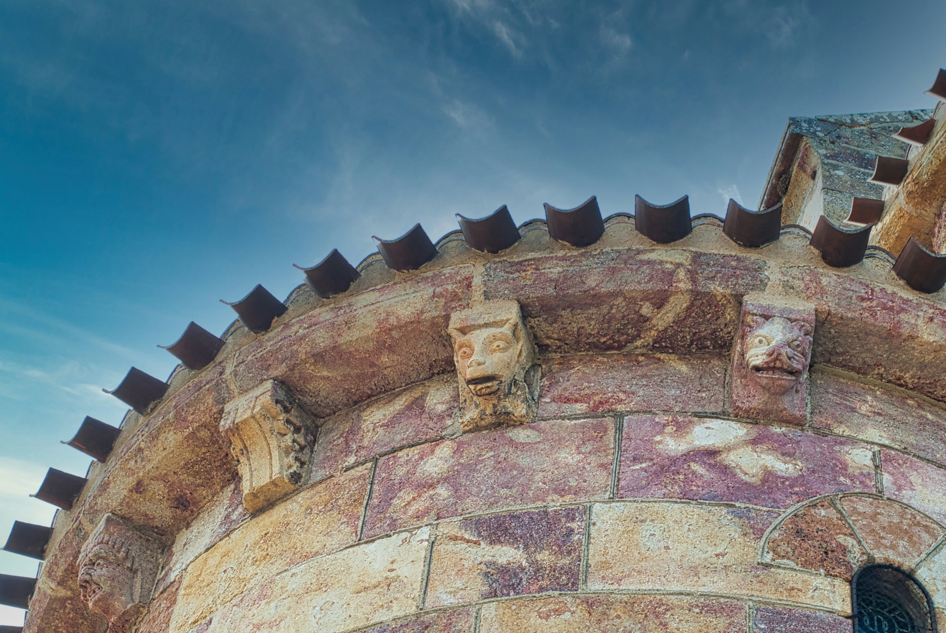 Curved stone wall with carved faces under a blue sky, showing architectural detail and decorative corbels.