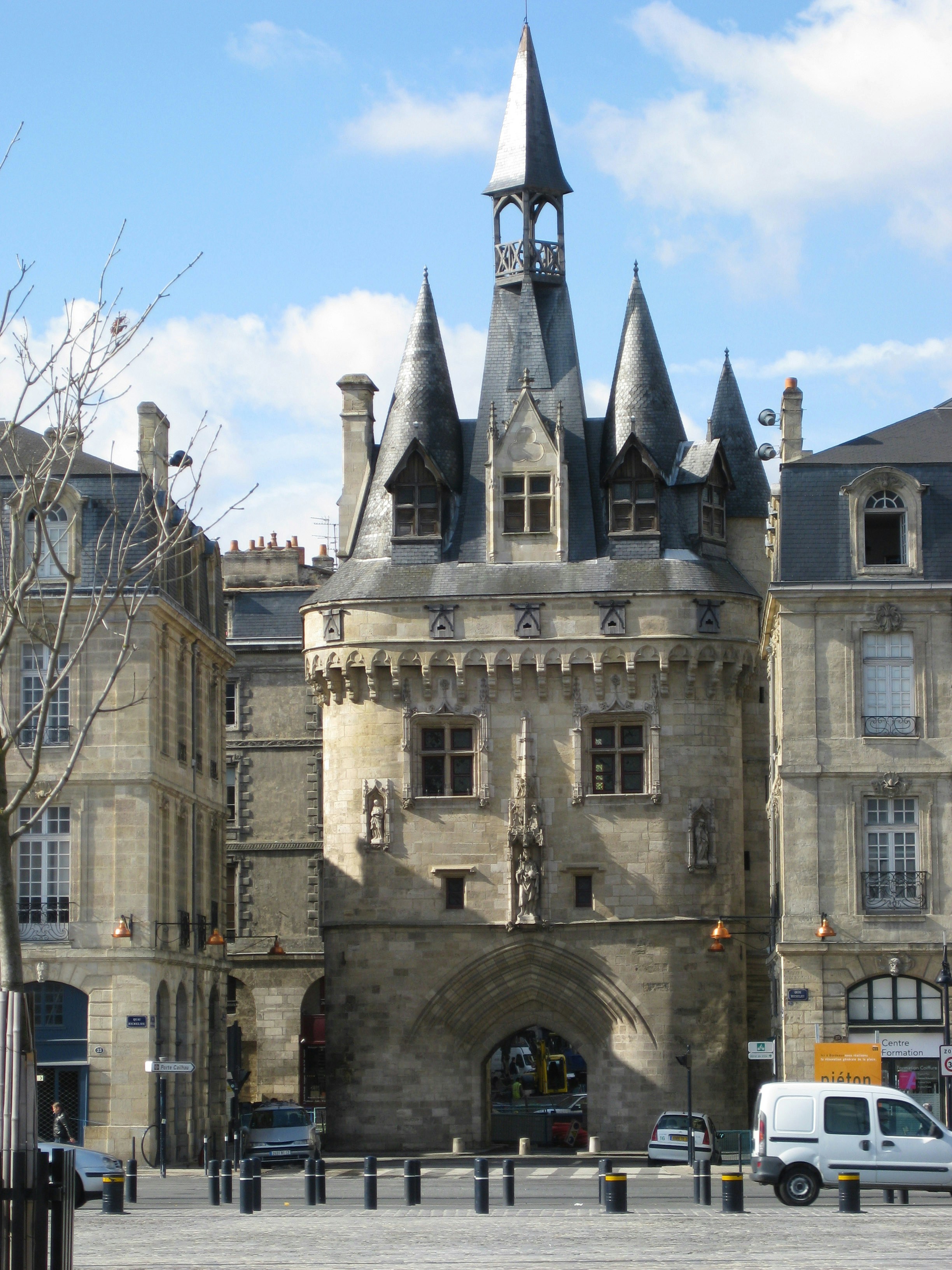 A historic stone gatehouse with conical towers stands as the focal point of a city street, flanked by adjacent buildings and a parked minivan; the structure serves as an architectural centerpiece in daylight.