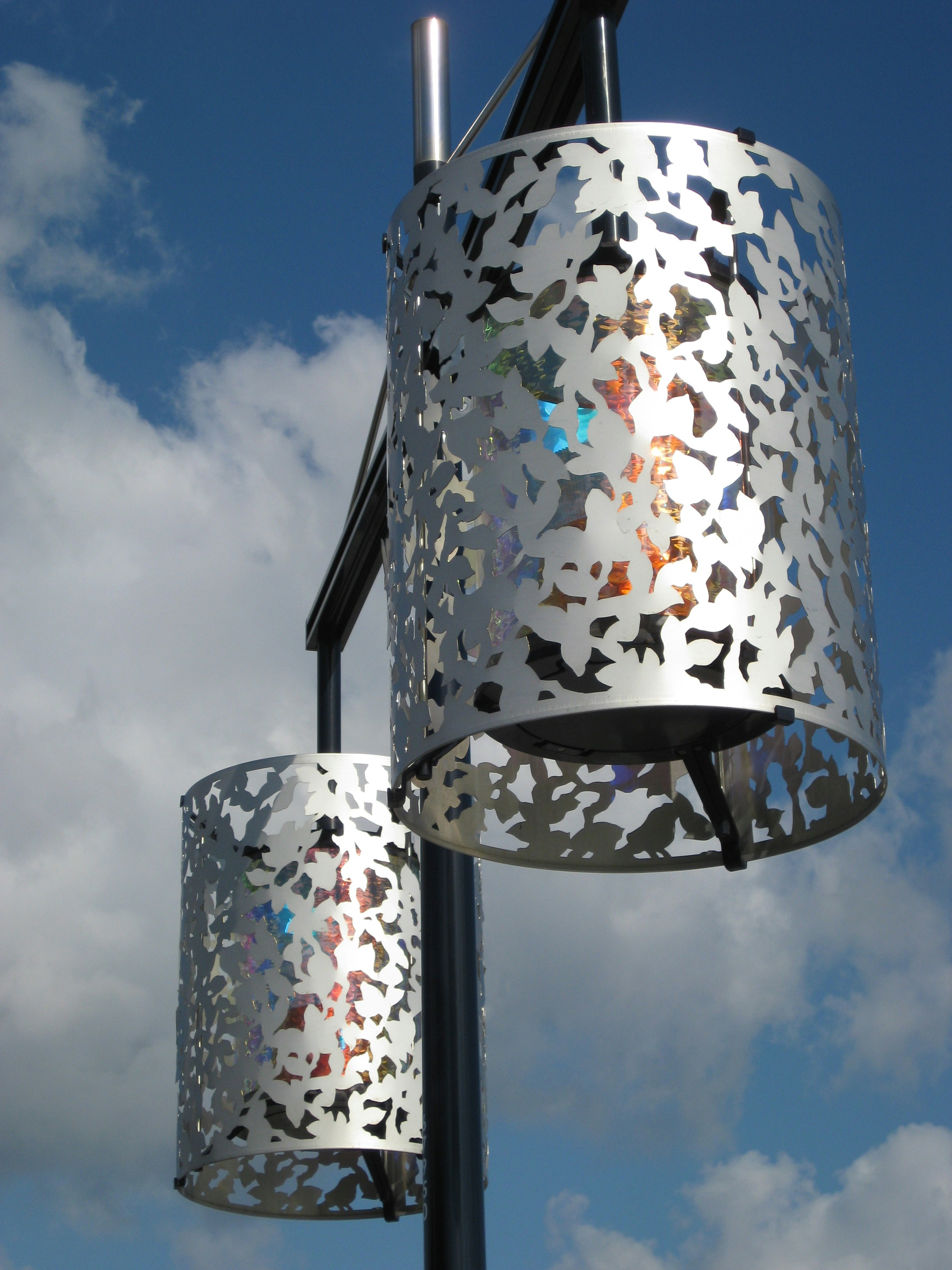 Two perforated metal lanterns hang from a vertical pole against a bright blue sky with clouds. The foreground lantern is large, metallic and sculpted with irregular cutouts that catch the sunlight, while a smaller lantern recedes in the background.