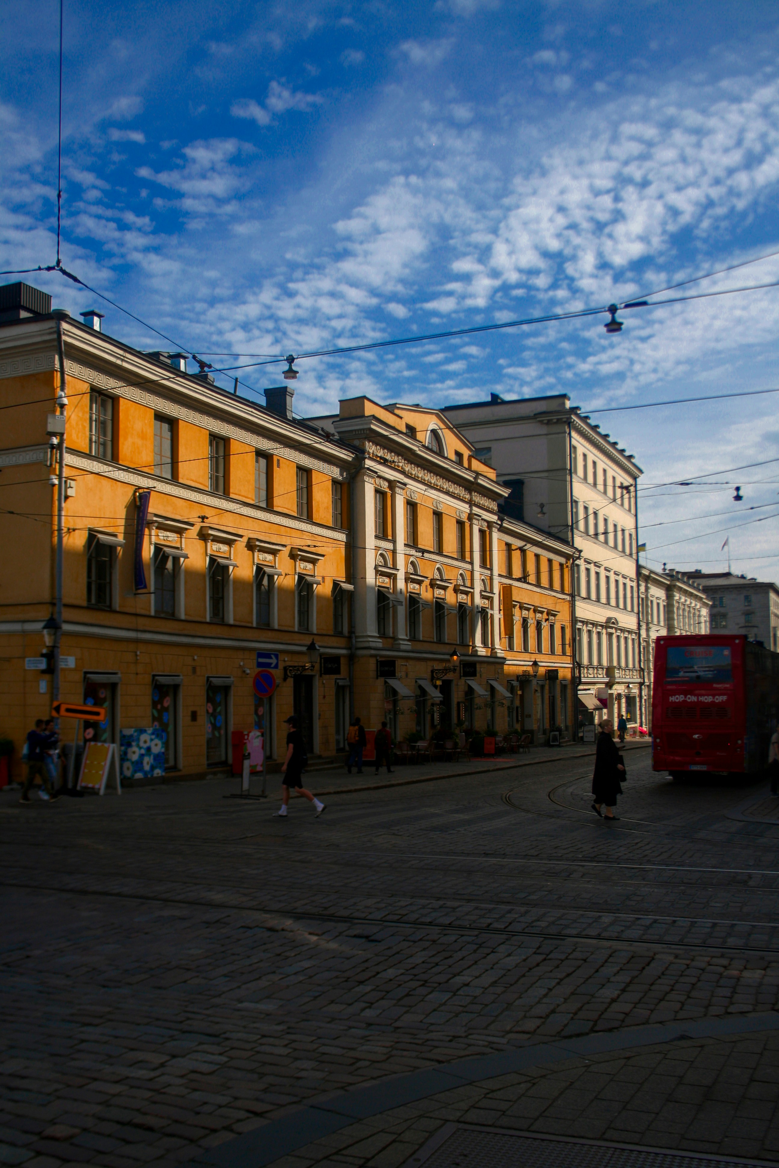 Warm-hued buildings line a cobblestone street as afternoon light bathes the façades; a red bus and pedestrians add urban rhythm to the scene. ( Photograph )