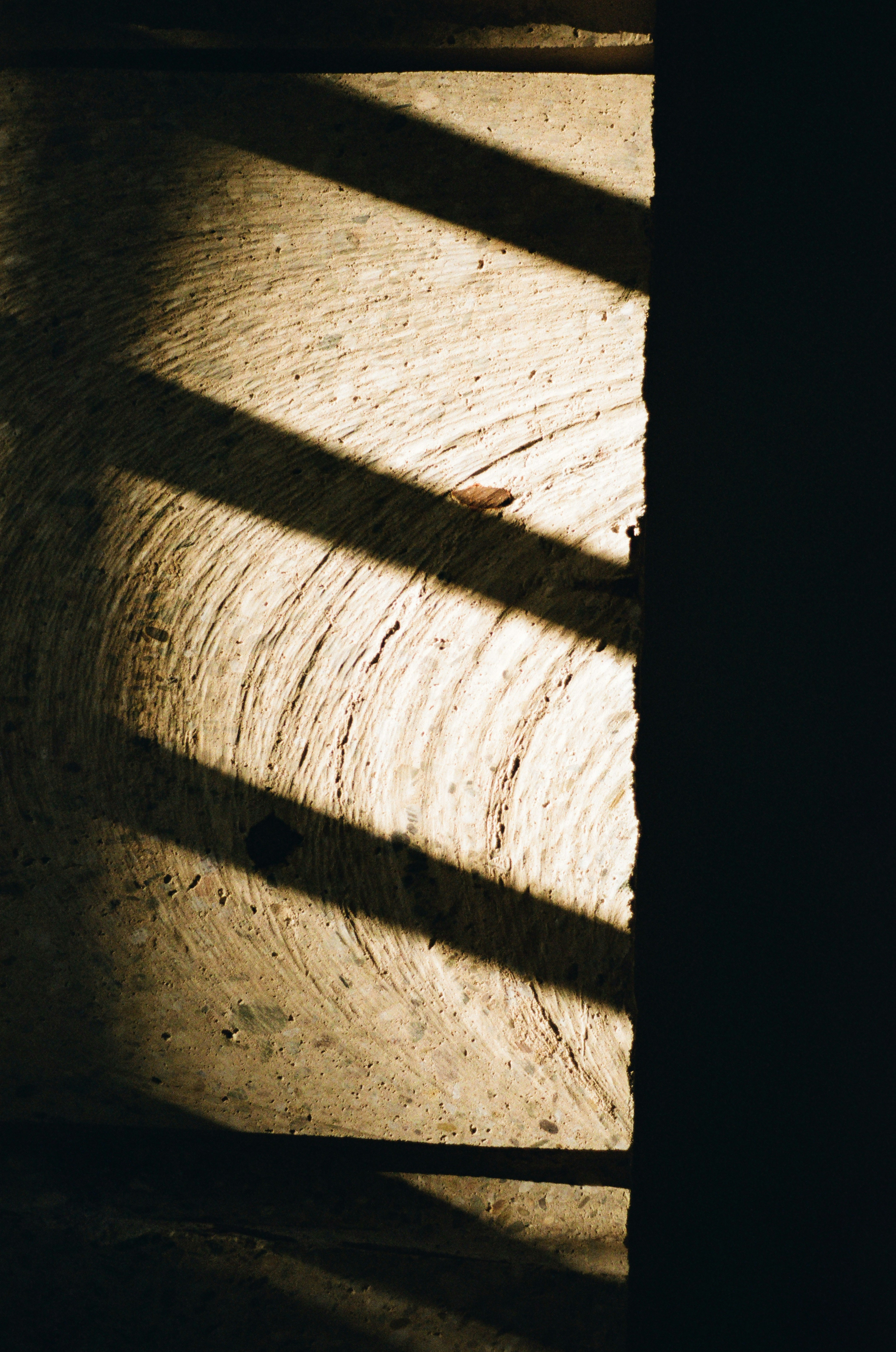 Concrete wall illuminated by angled light bands, creating bold shadow patterns across the surface. A stark vertical dark edge frames the right side, emphasizing texture and composition.