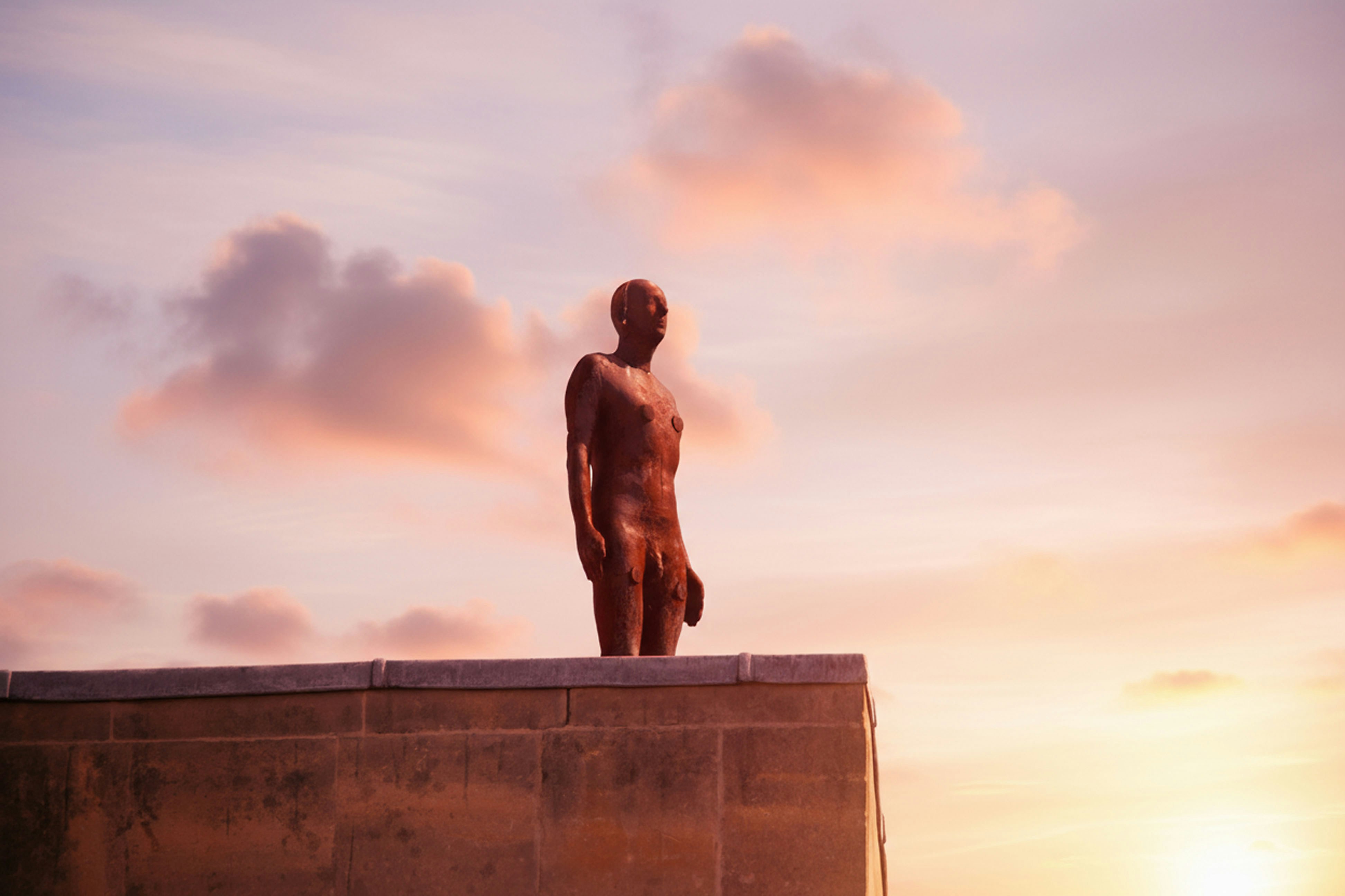 Bronze statue atop a concrete ledge against a warm sunset sky; the serene figure becomes the focal point as clouds glow softly in pastel hues. Sculpture emphasizes calm, minimal surroundings.