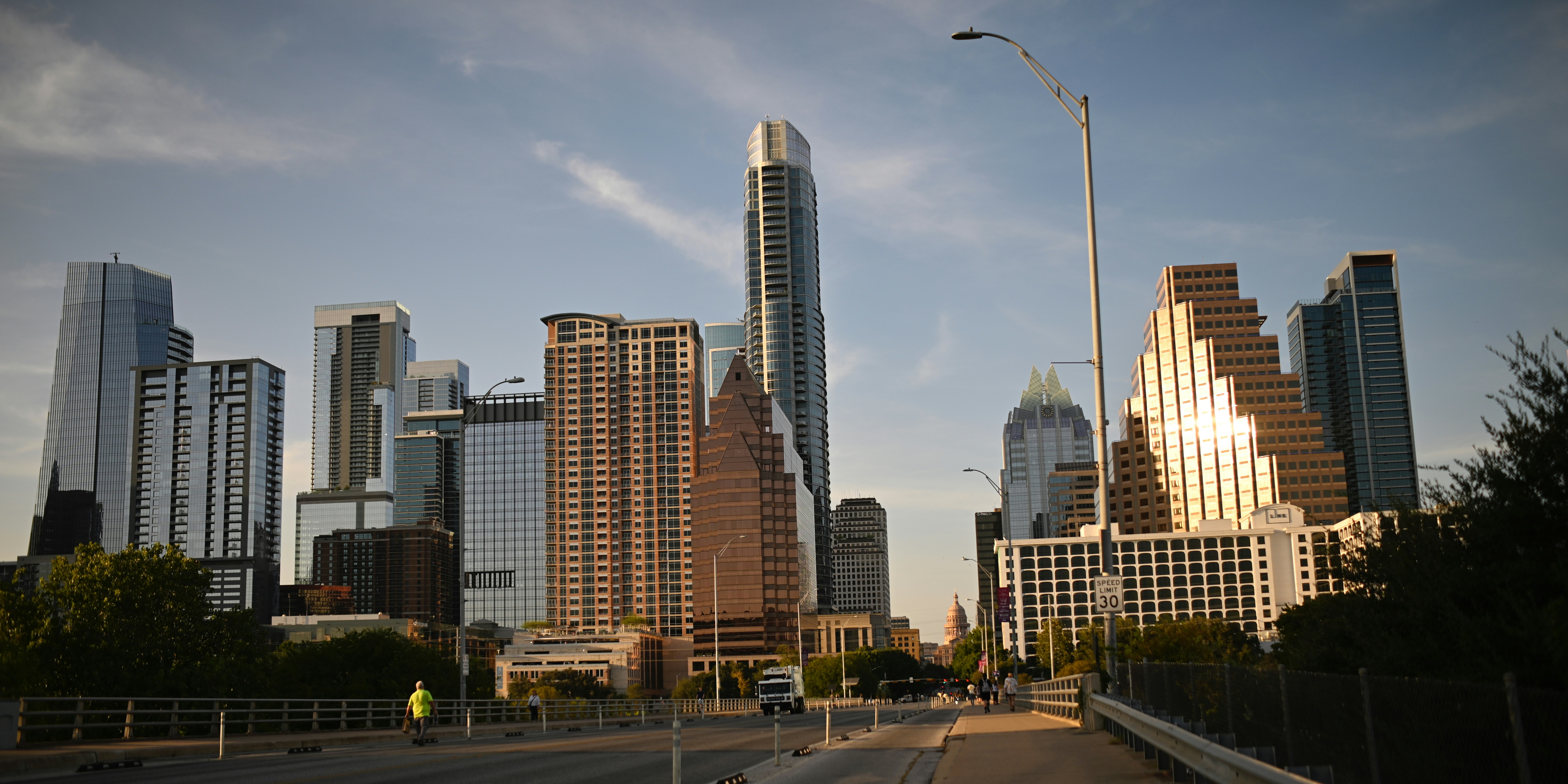 City skyline with modern high-rrise towers rising above a riverfront promenade and trees; a quiet, late-afternoon view capturing reflective glass and stacked architecture.