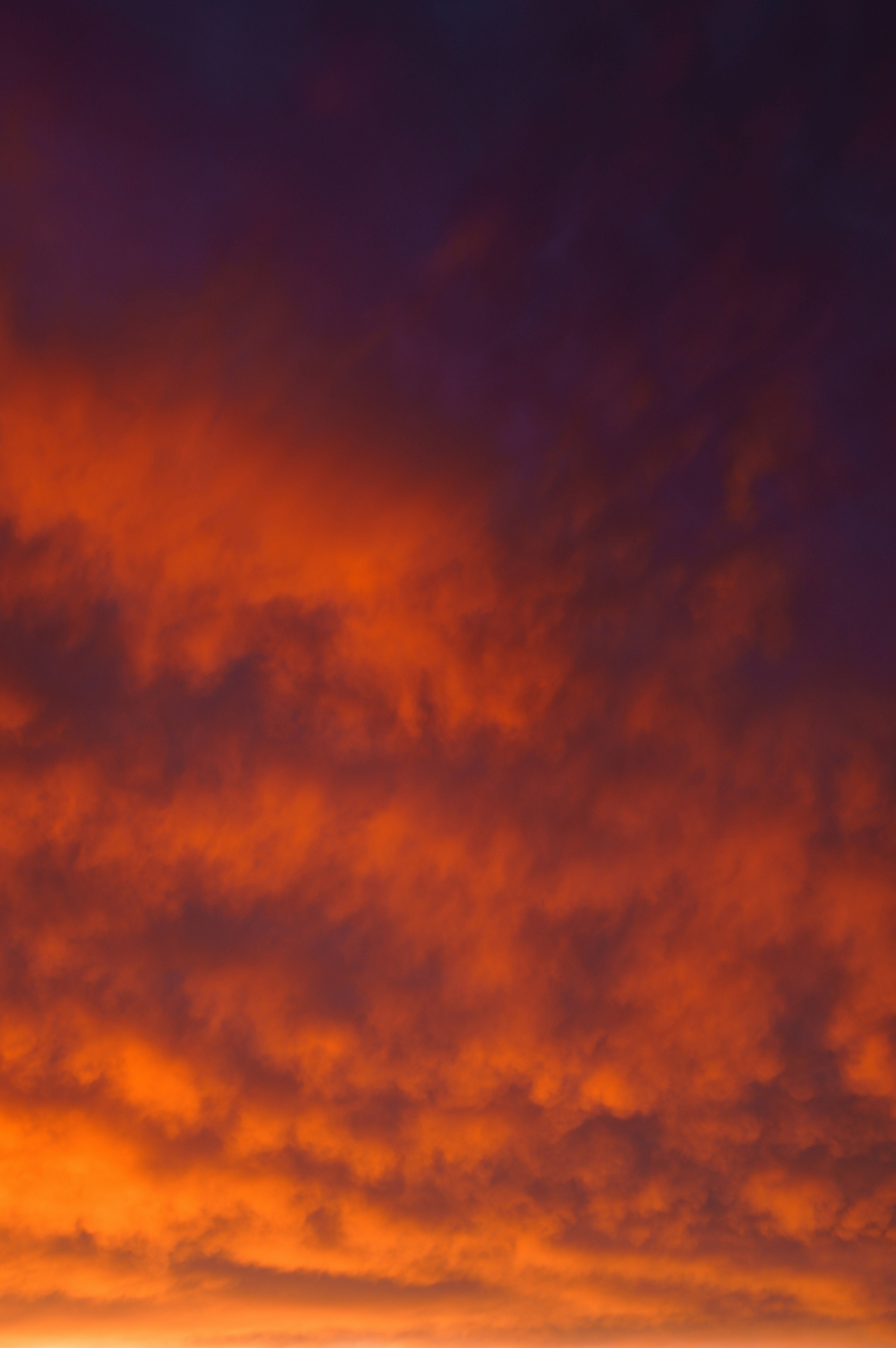 Dramatic orange clouds lit by a setting sun sweep across a darkening sky, creating a bold gradient above a calm horizon. The scene emphasizes color and texture of the clouds.