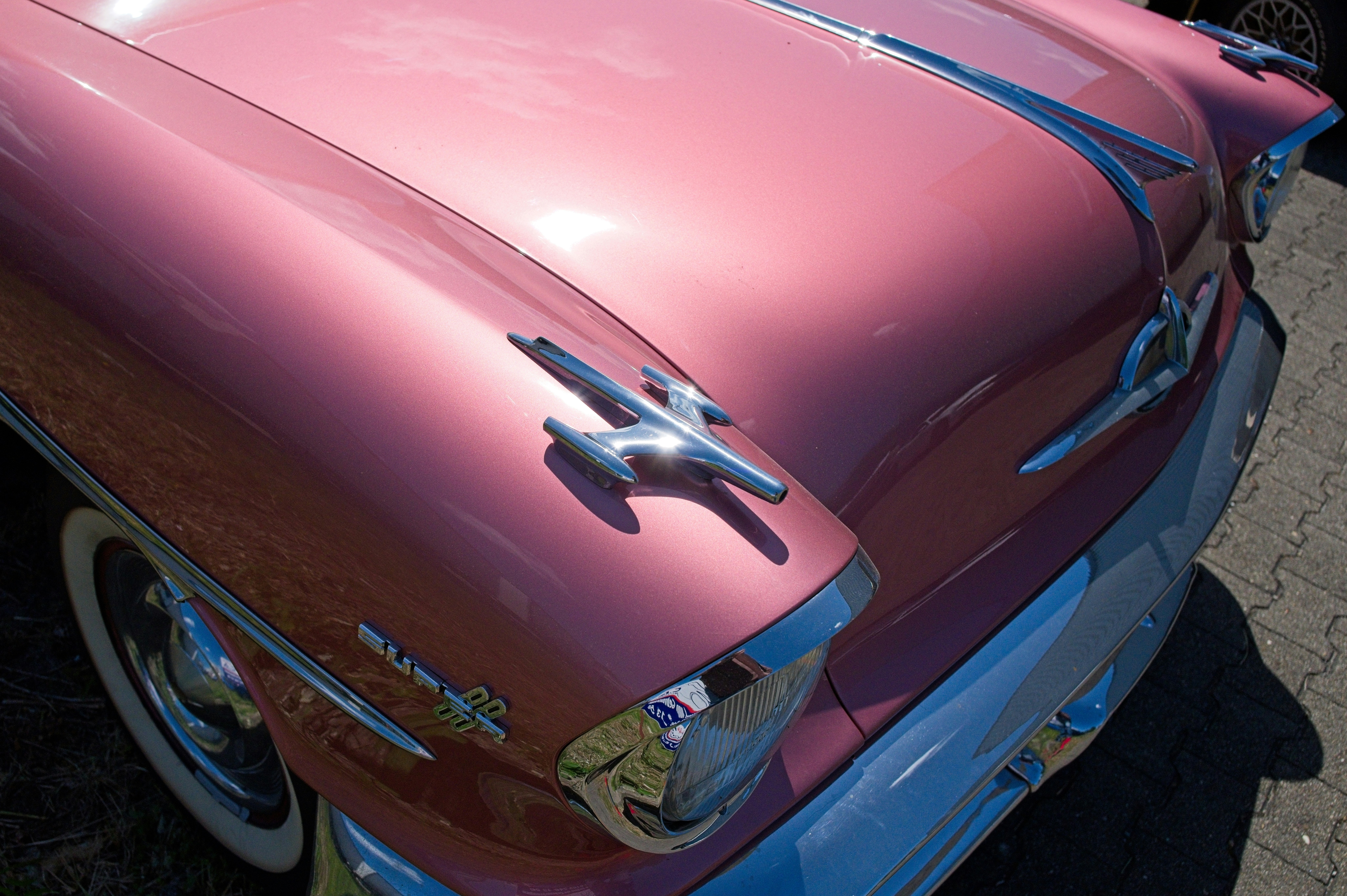 Glossy pink vintage car hood with chrome emblem and emblematic badge, reflecting surroundings; image emphasizes metallic paint, chrome trim, and retro lines of a 1950s Chevrolet.