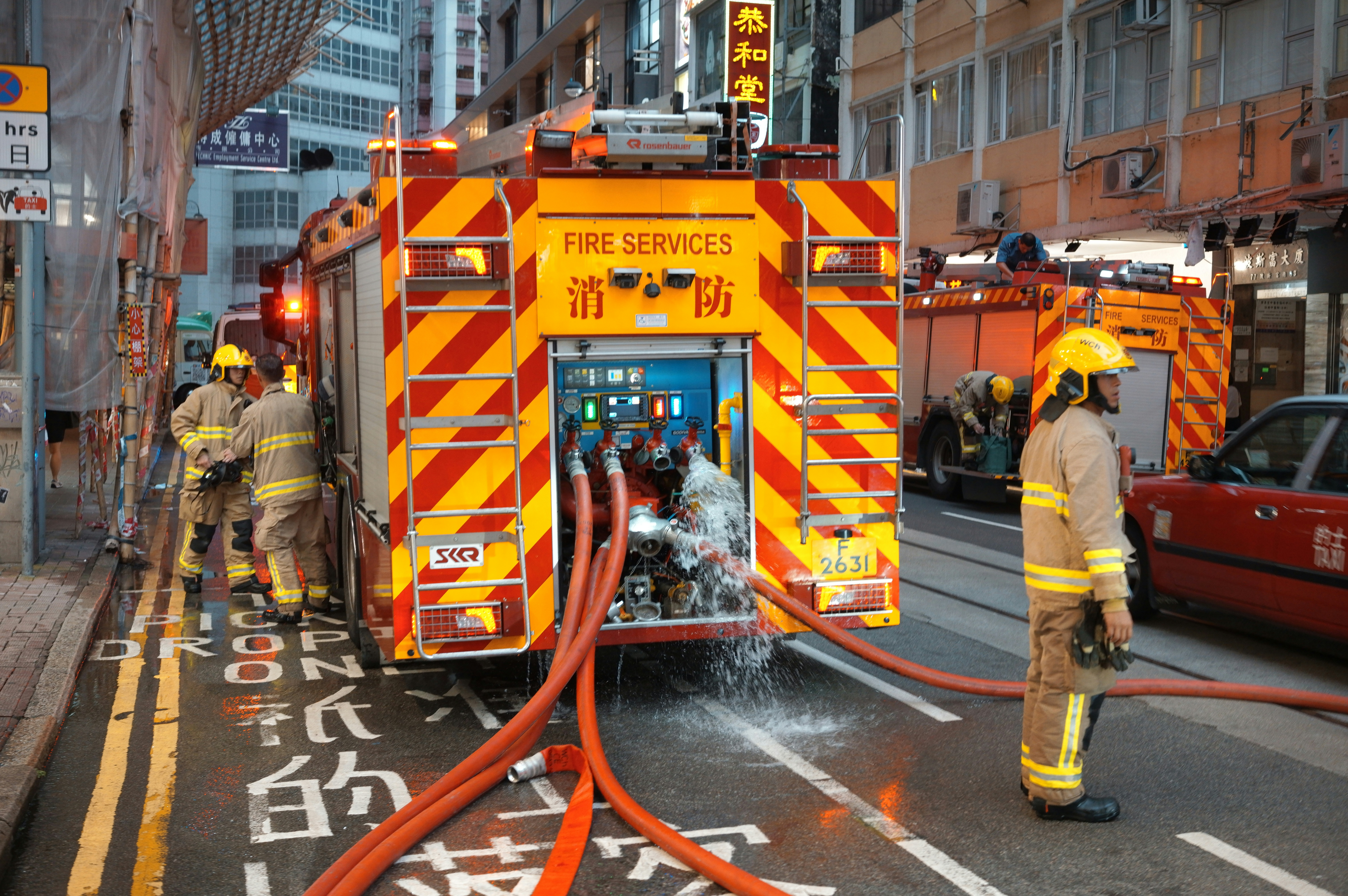 Firefighters work beside a bright orange fire engine with hoses spraying water on a city street.