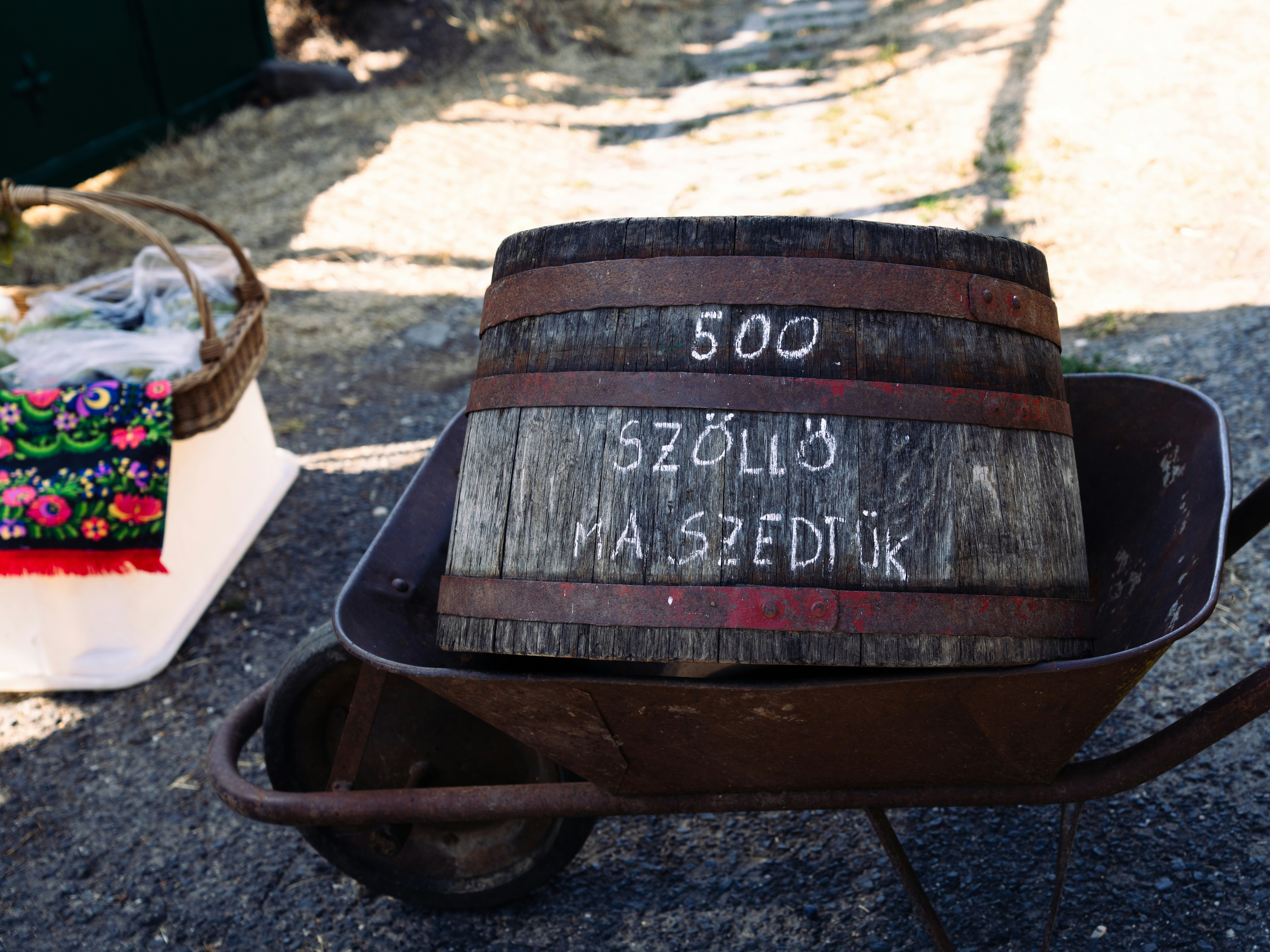Barrel with chalked markings sits atop a rustic wheelbarrow in a sunlit outdoor setting, hinting at a market or rustic display.