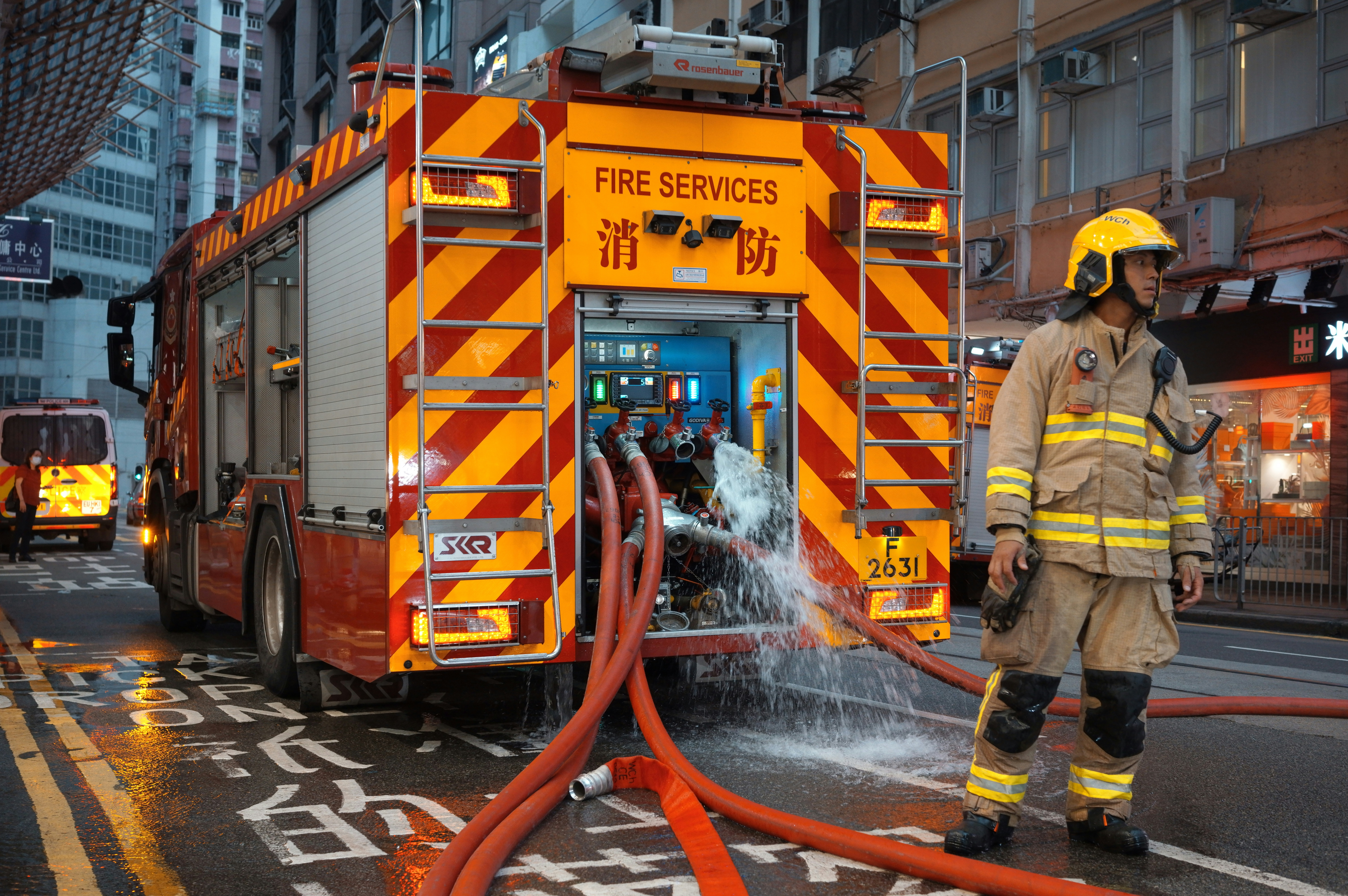 Fire engine spraying water hoses in a city street with a firefighter standing nearby; wet pavement and urban storefronts frame the scene.