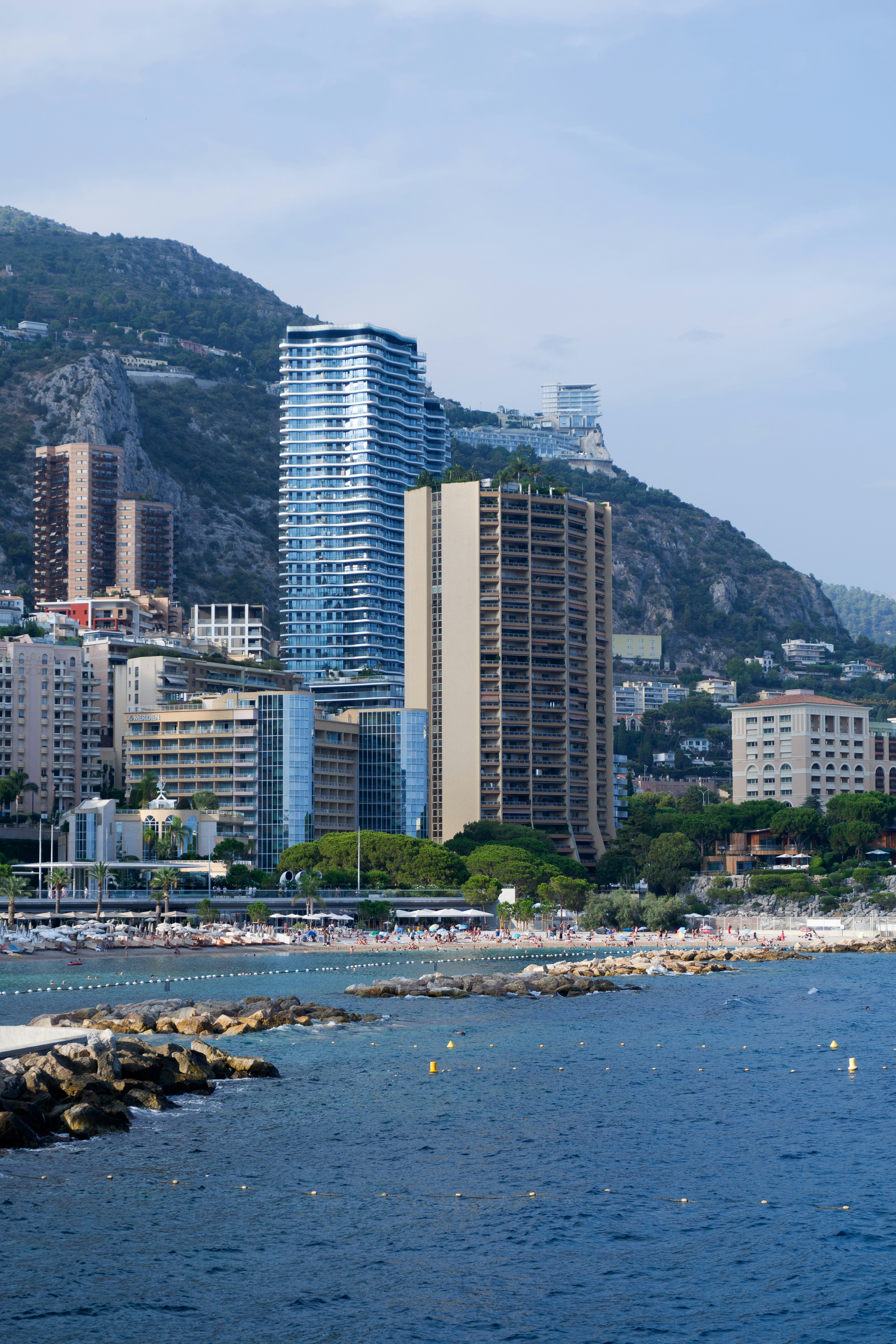 Coastal cityscape along a calm sea with high-rise buildings lining the shore and a rocky breakwater in the foreground.