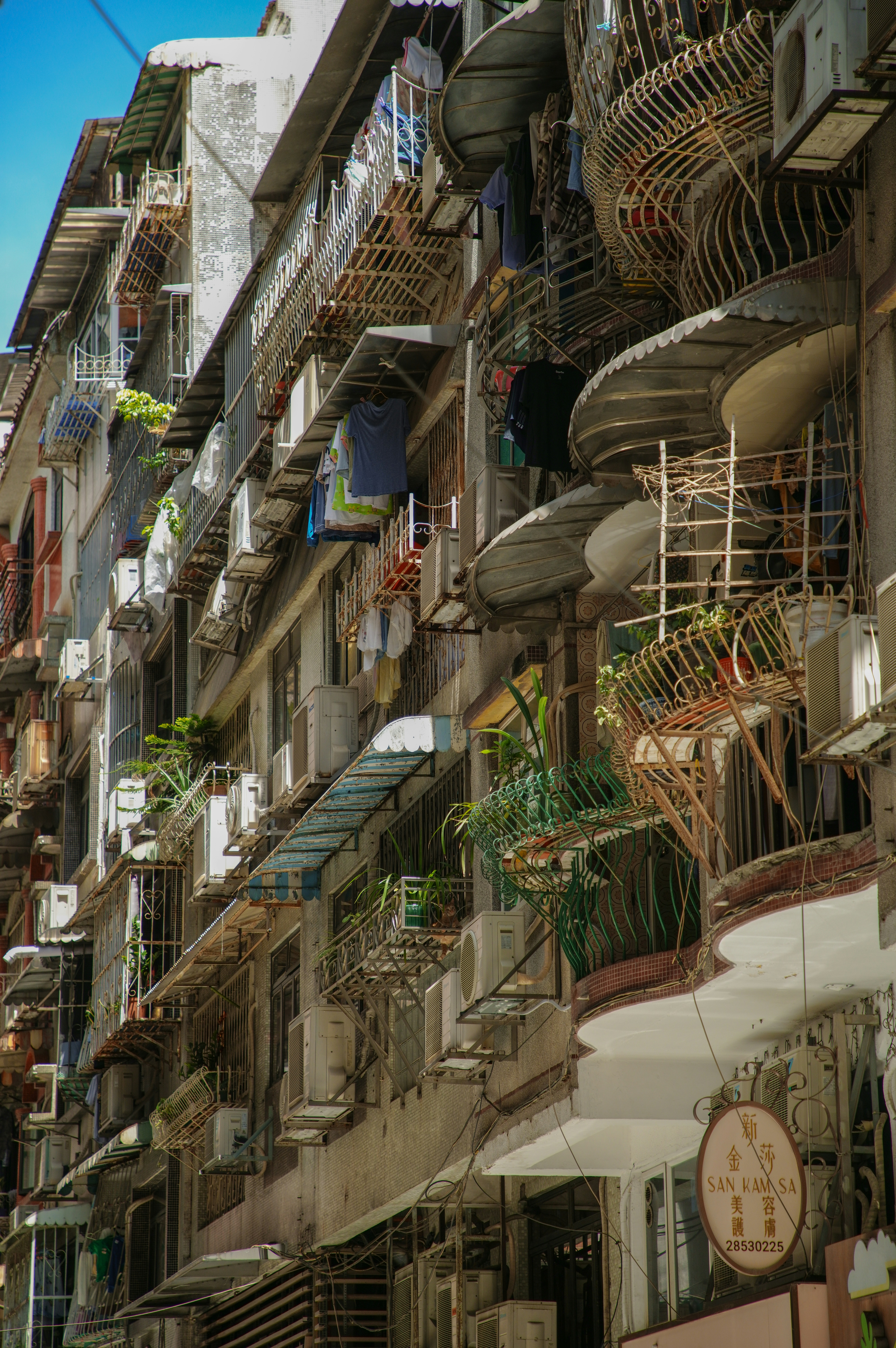 Dense urban facade with stacked balconies, window units, and metal grilles; sunlight reveals varied textures and plants on the building fronts.