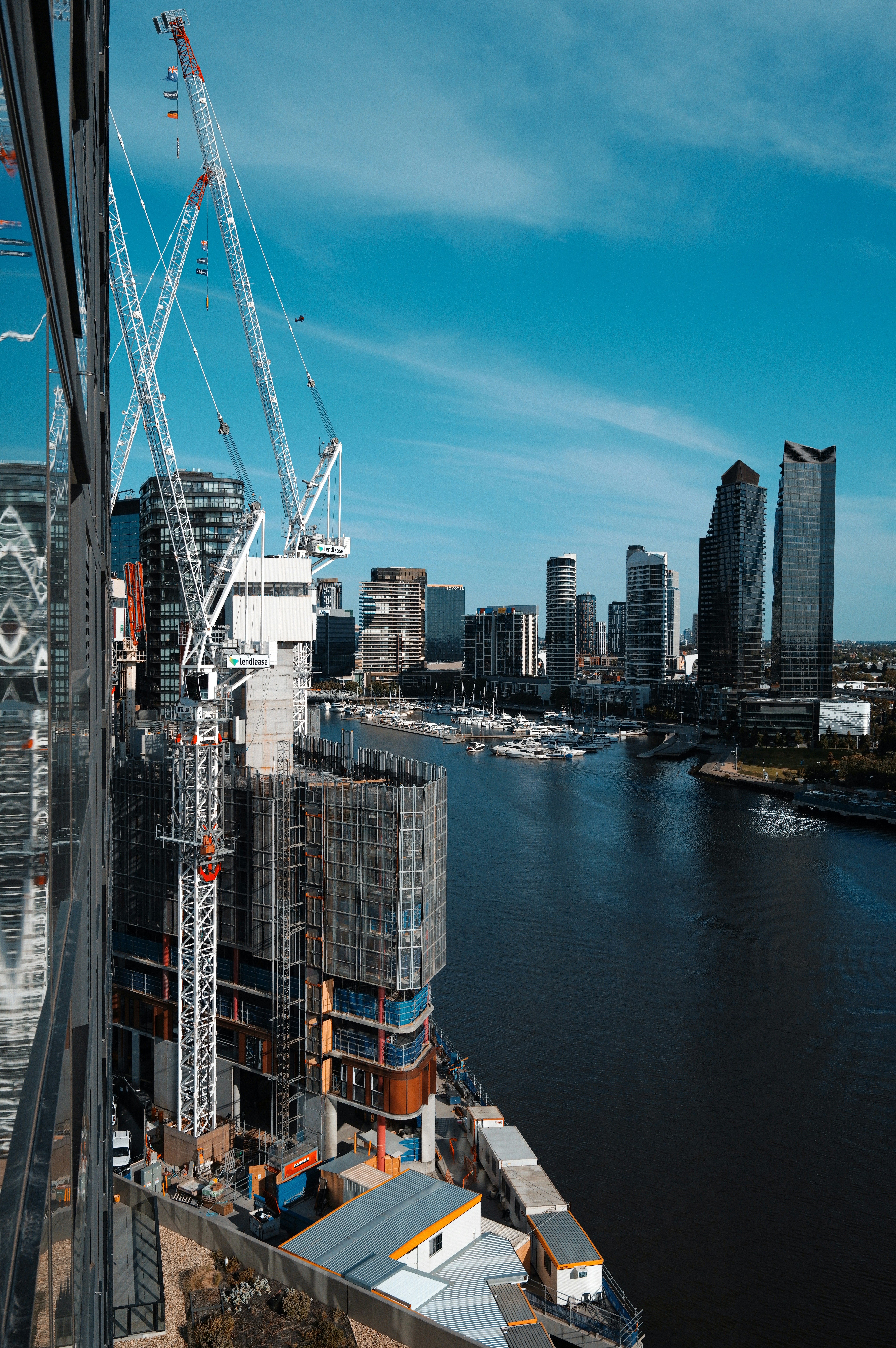 Construction cranes rise beside a riverfront with modern towers stretching into a blue sky.