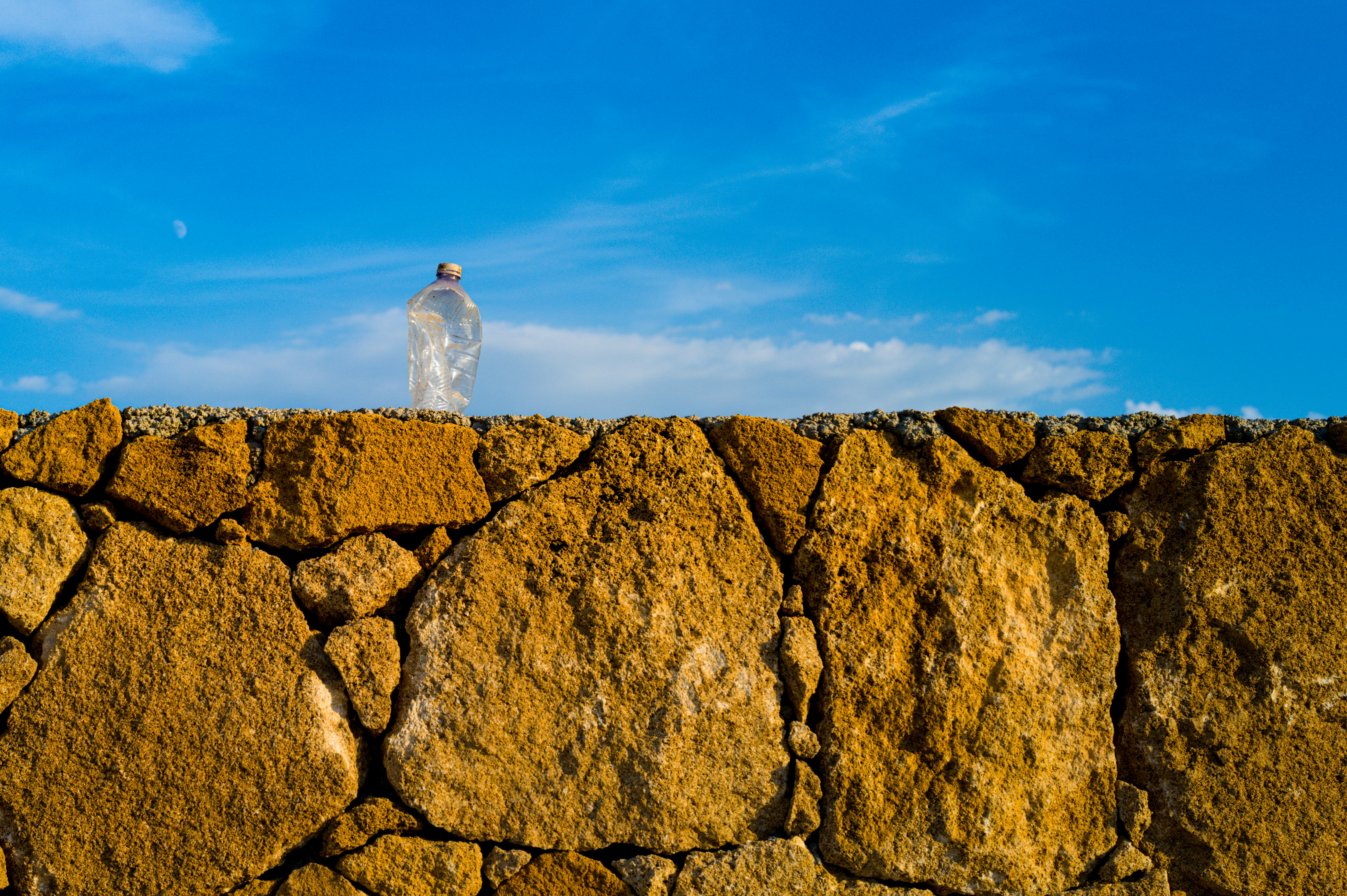 Clear, sunlit brick wall with a lone plastic bottle near the top above the wall, set against a vibrant blue sky.