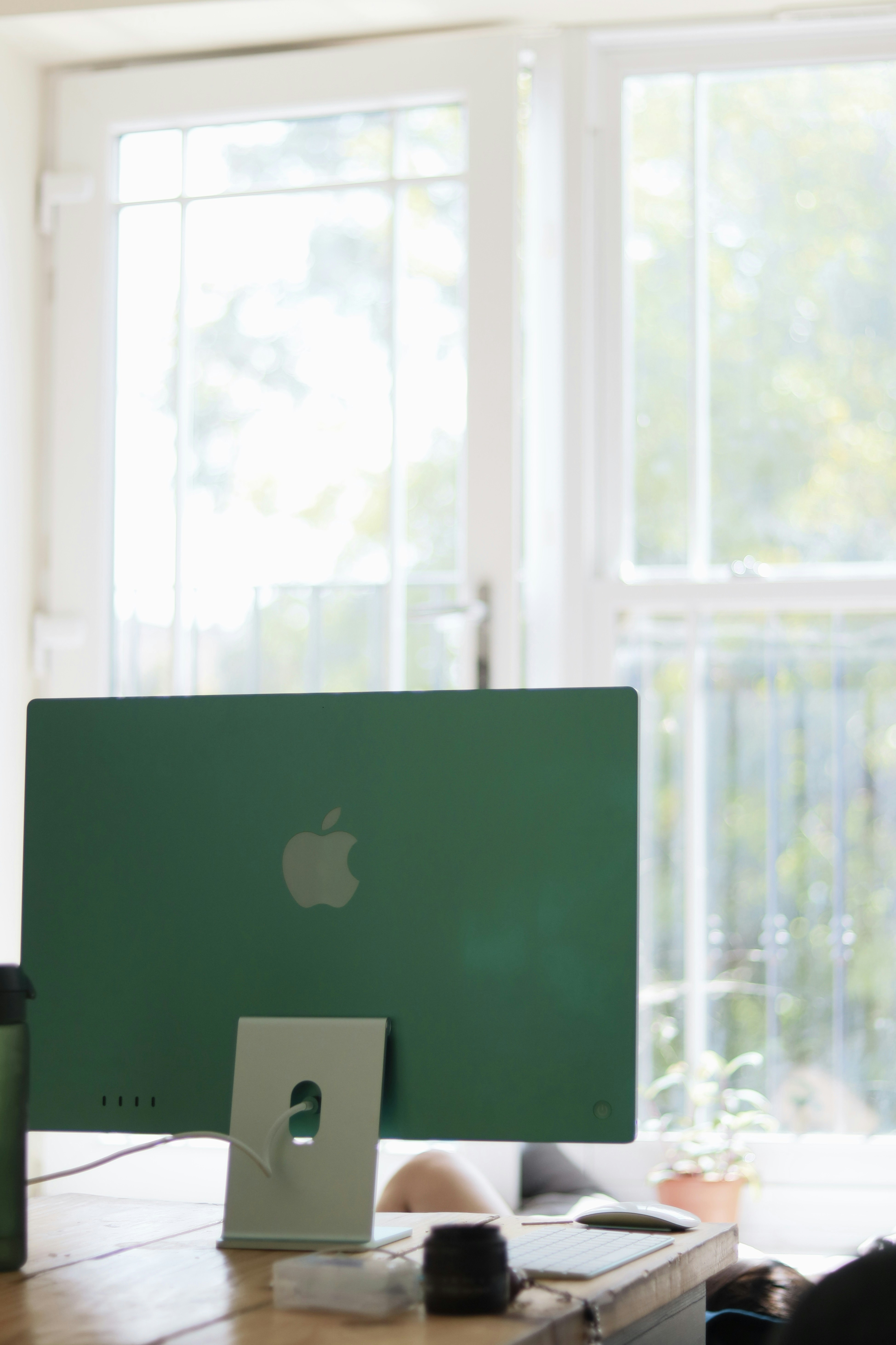 Mint-green iMac on a wooden desk by large windows with daylight; minimalist workspace and small plant pot in background.