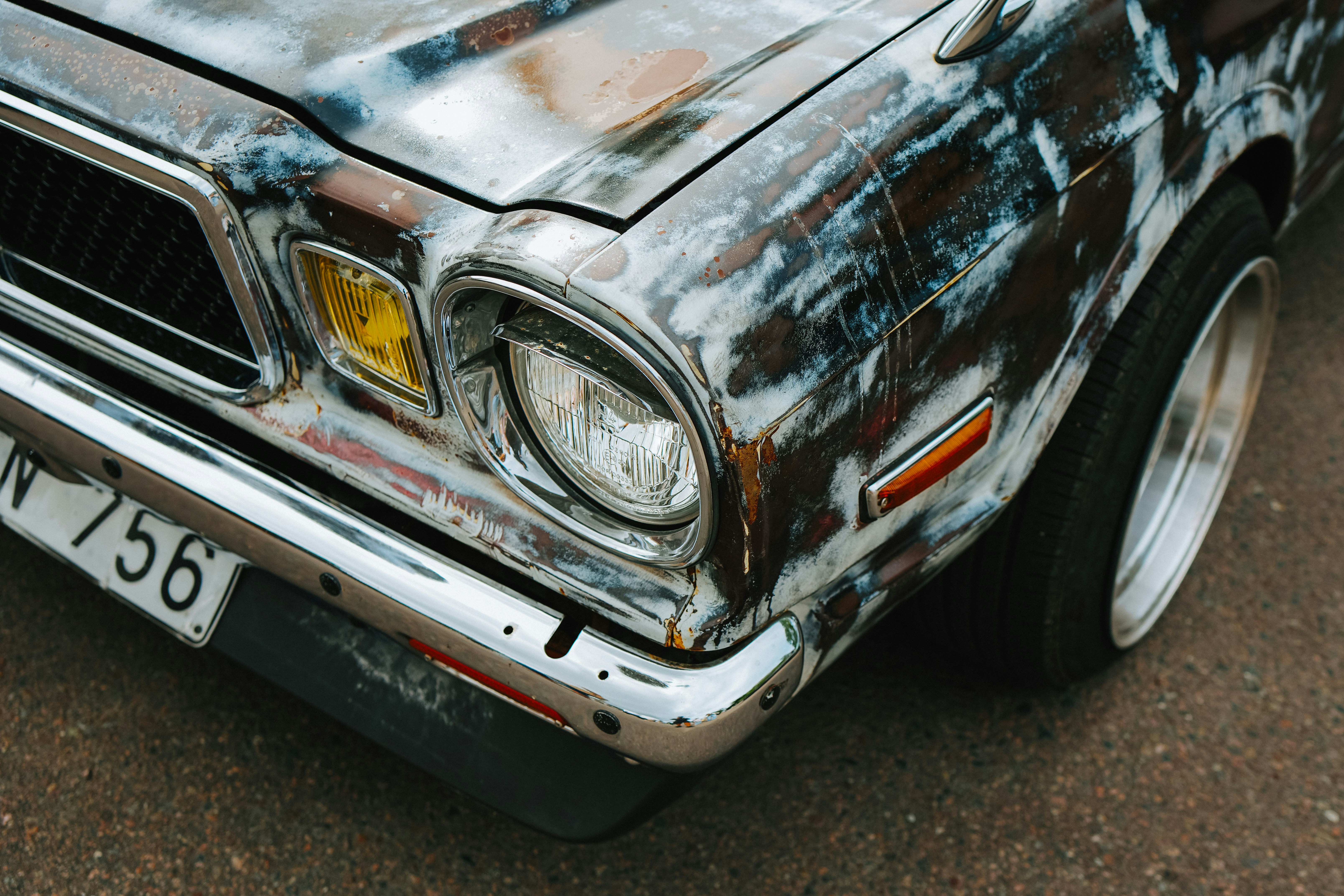 The front corner of a weathered car with rusted paint and chrome bumper