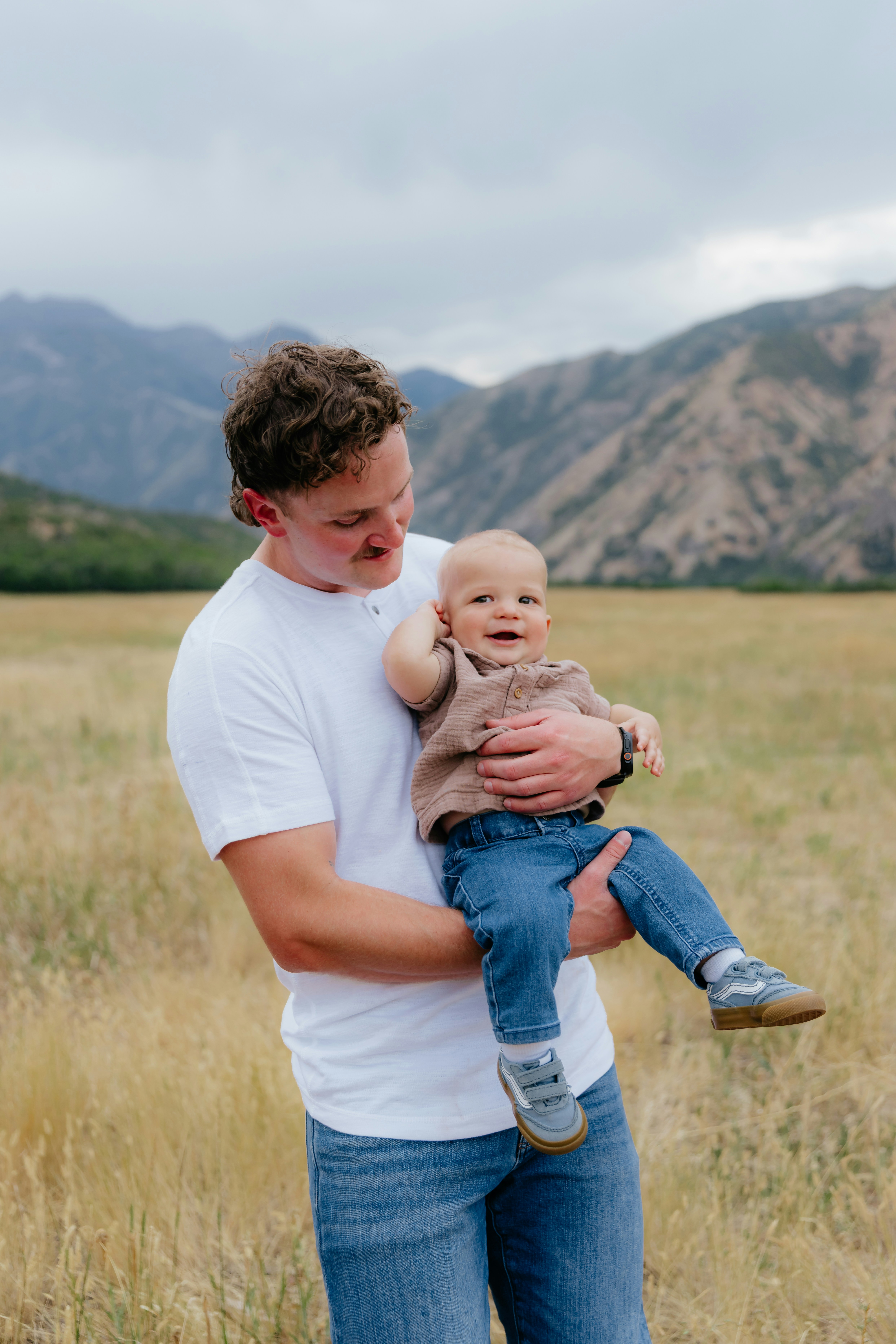 Father in White T-shirt Lifts Baby Against Mountain Field