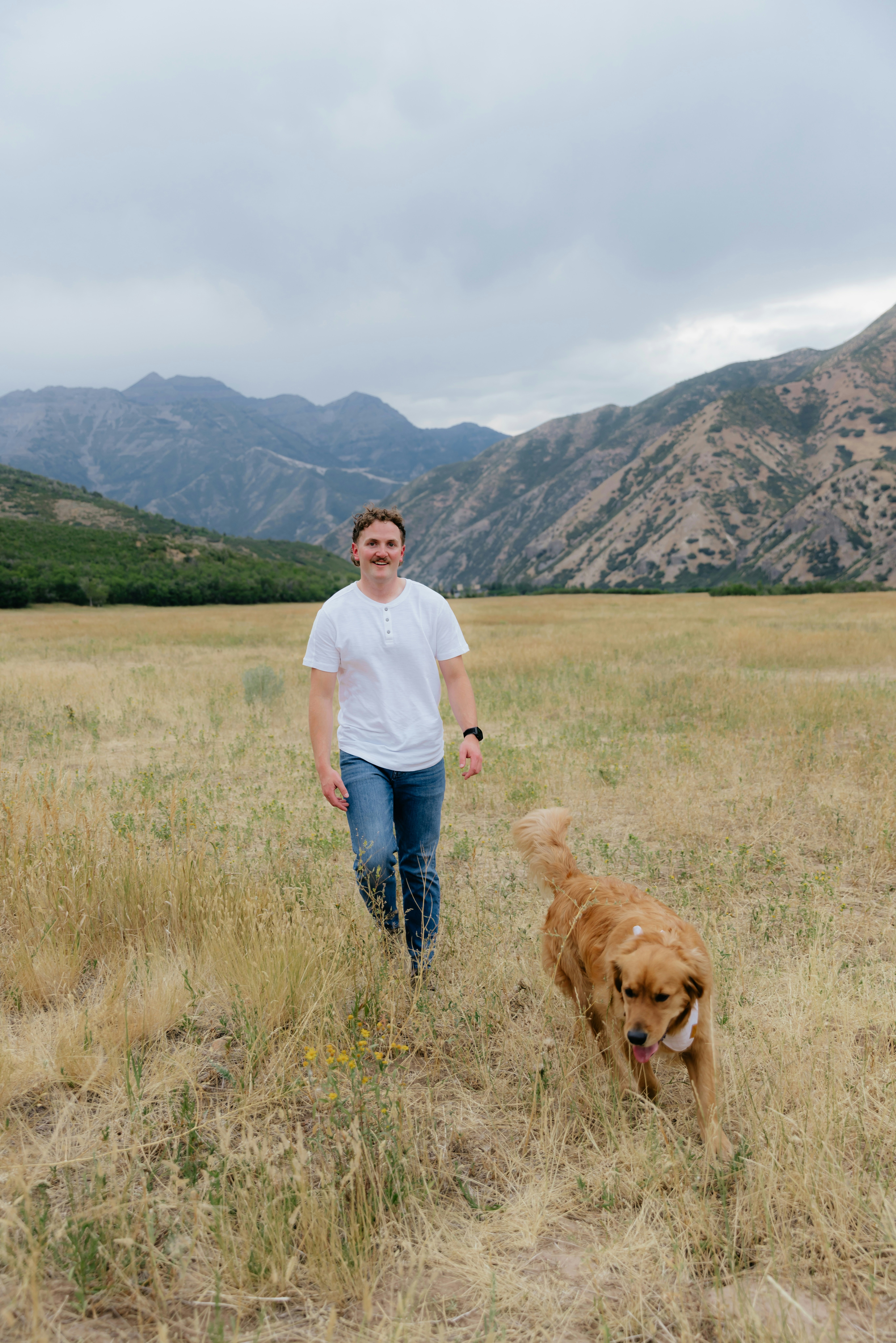 Man in white tee walking in golden meadow with dog as mountains loom