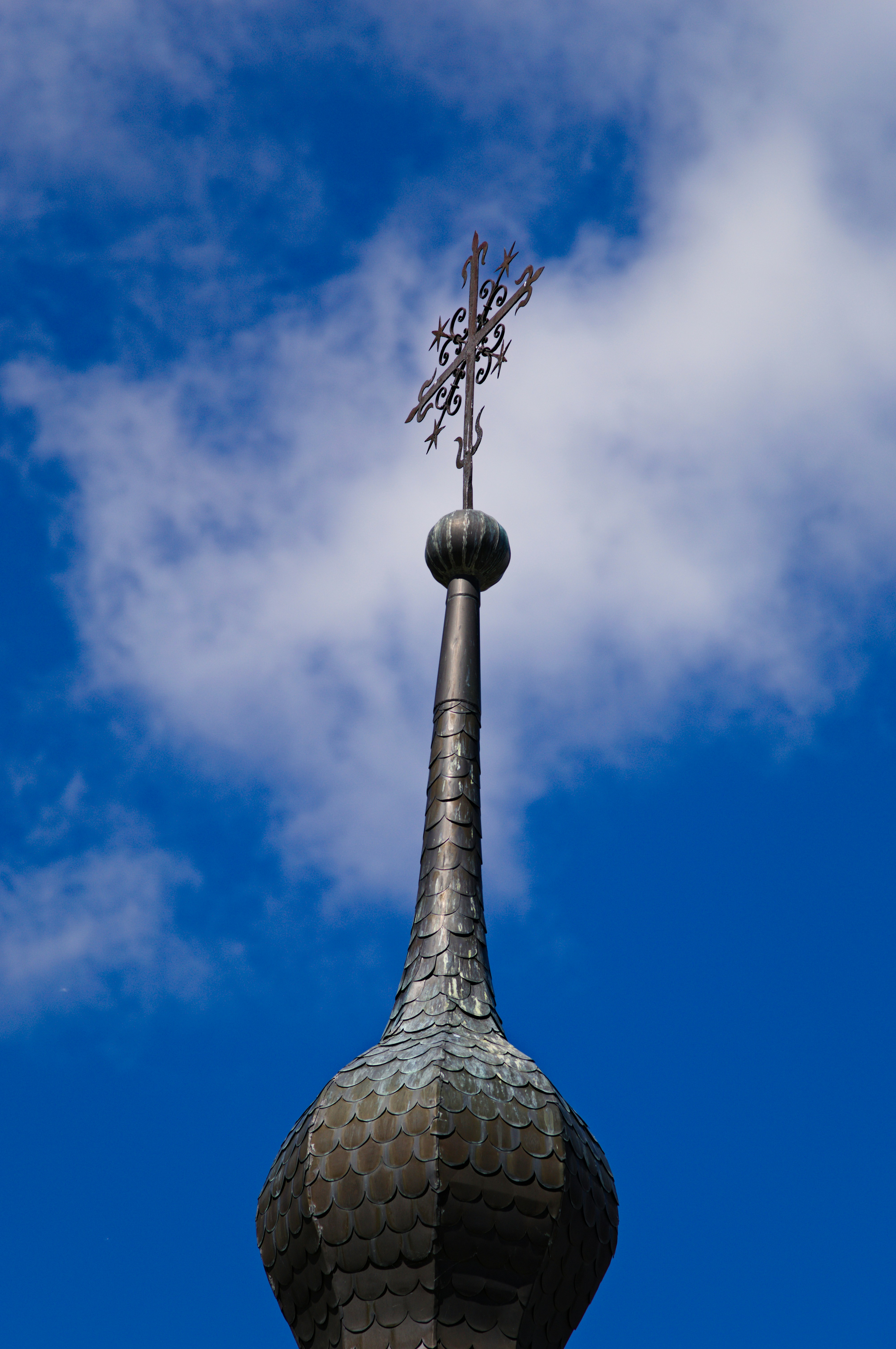 Metal spire with ornate cross on top rises against a blue sky, captured with strong vertical composition and sharp scalloped roofing texture.