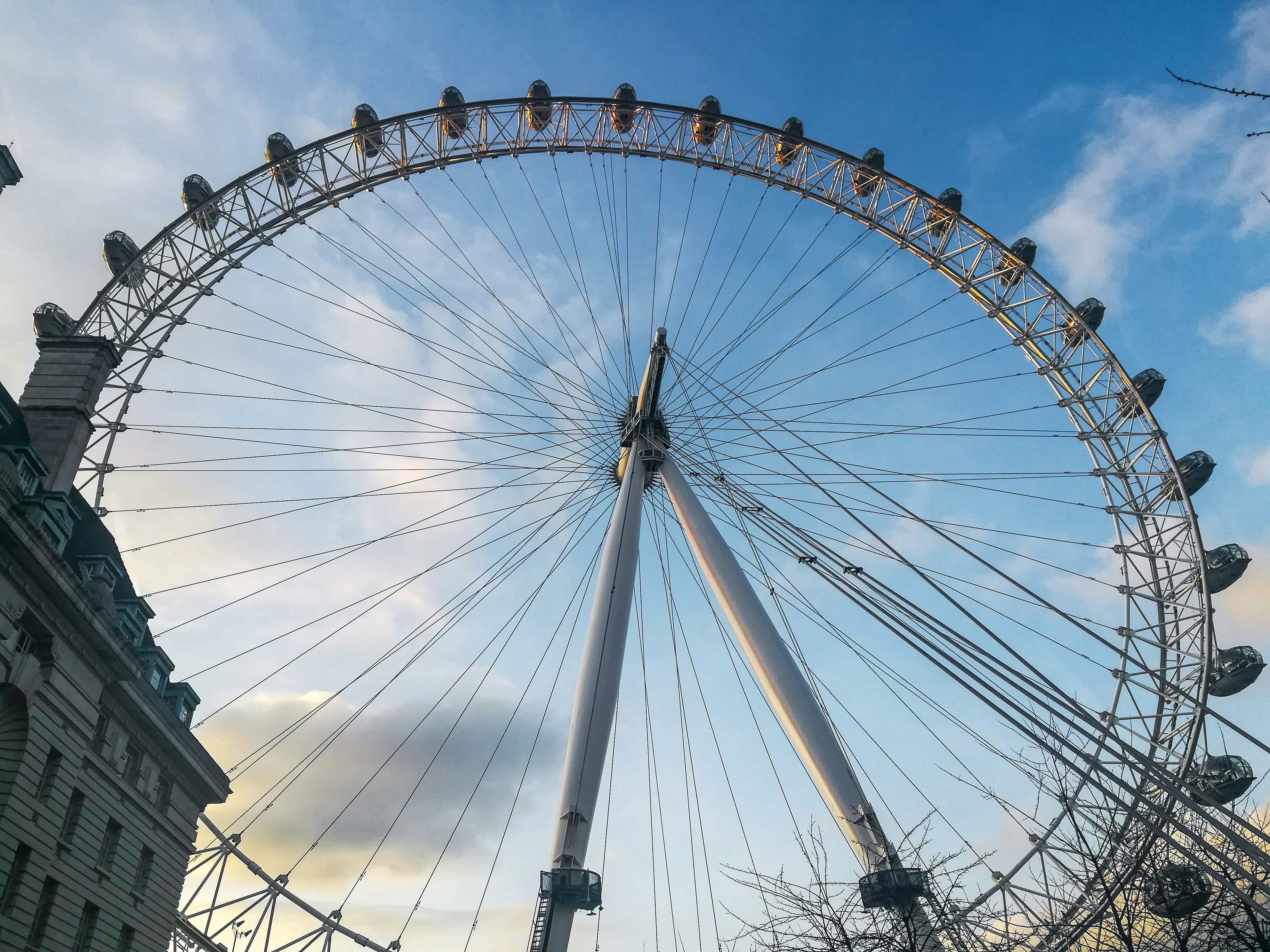 Close-up view of the London Eye ferris wheel framed against a blue sky with scattered clouds; structural spokes and supporting hub are clearly visible, giving a sense of scale and engineering.