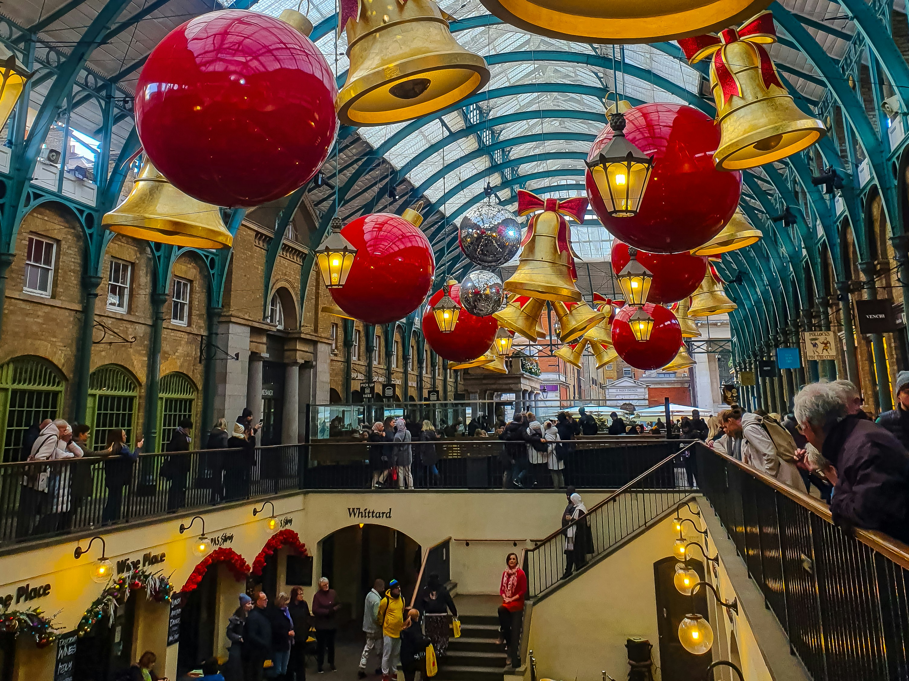 Decorated indoor market with oversized red and gold holiday ornaments hanging from a glass ceiling, shoppers moving along balconies and stairs. A festive scene in a retail arcade, showing architectural arches and warm lighting.