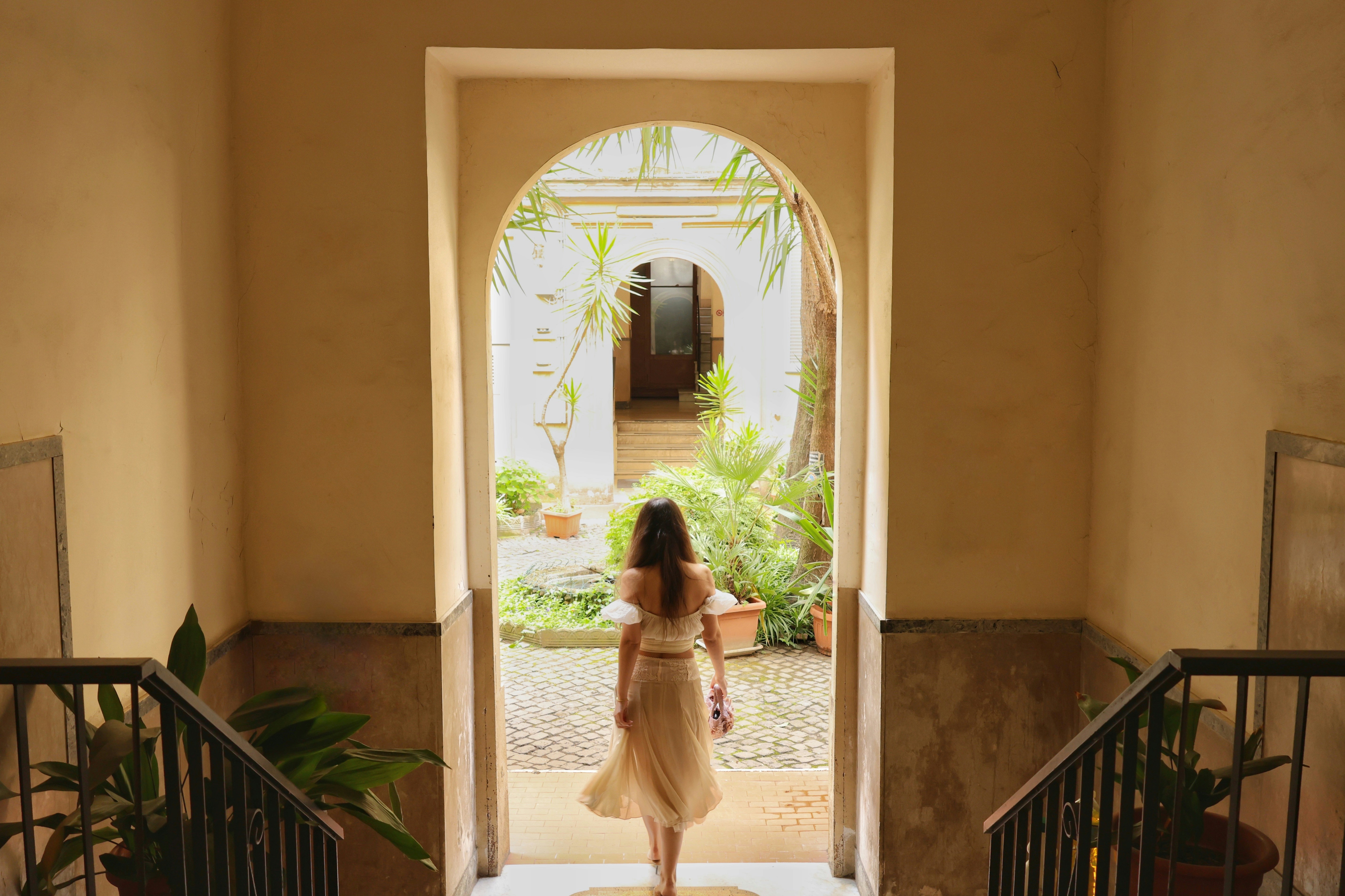 A woman in a flowing dress stands at an arched doorway, stepping into a sunlit courtyard framed by plants and stone walls.
