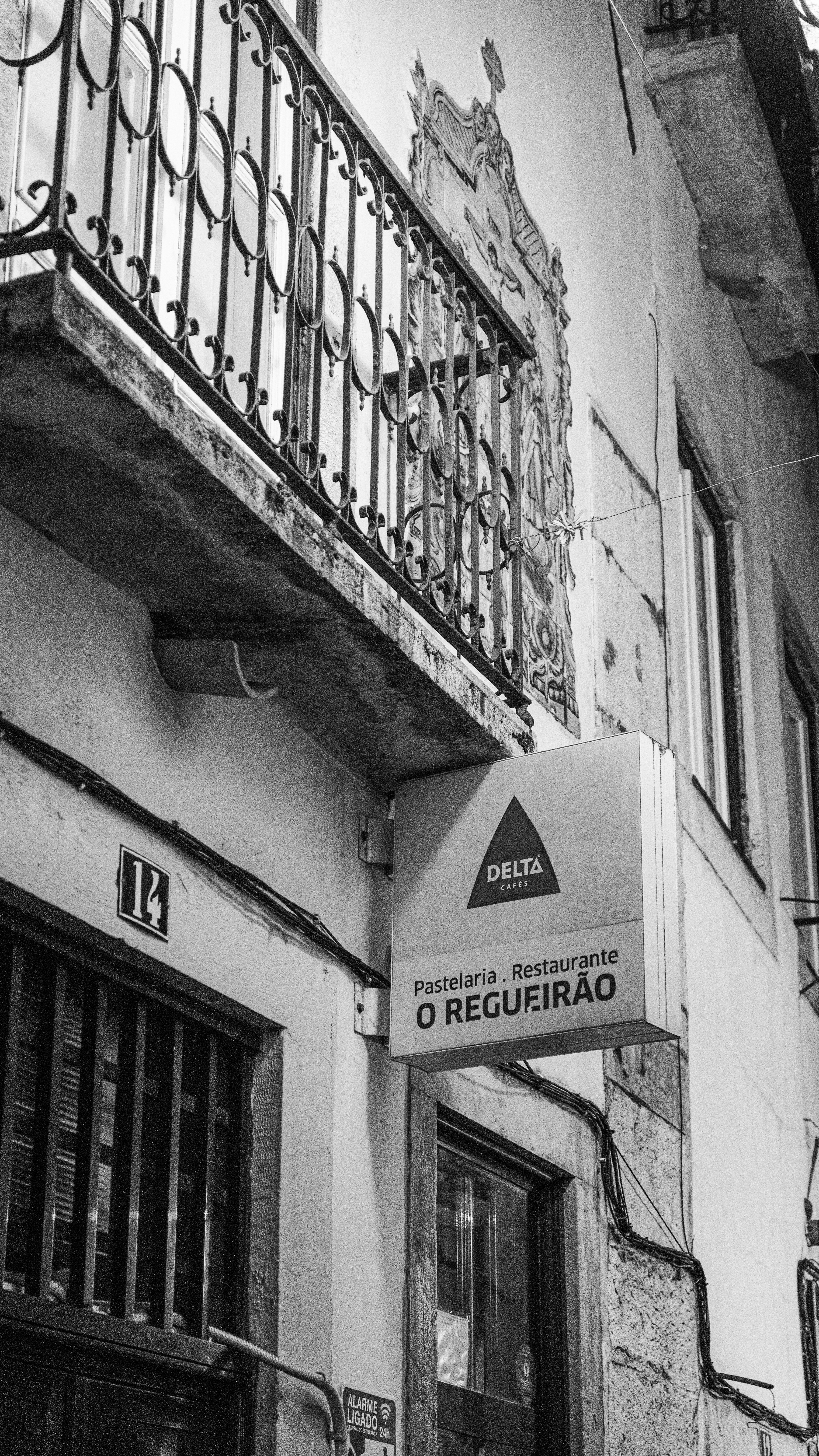 Black-and-white street scene featuring a corner bakery sign 'O REGUEIRÃO' beneath an ornate balcony, with architectural details and a doorway to the left.