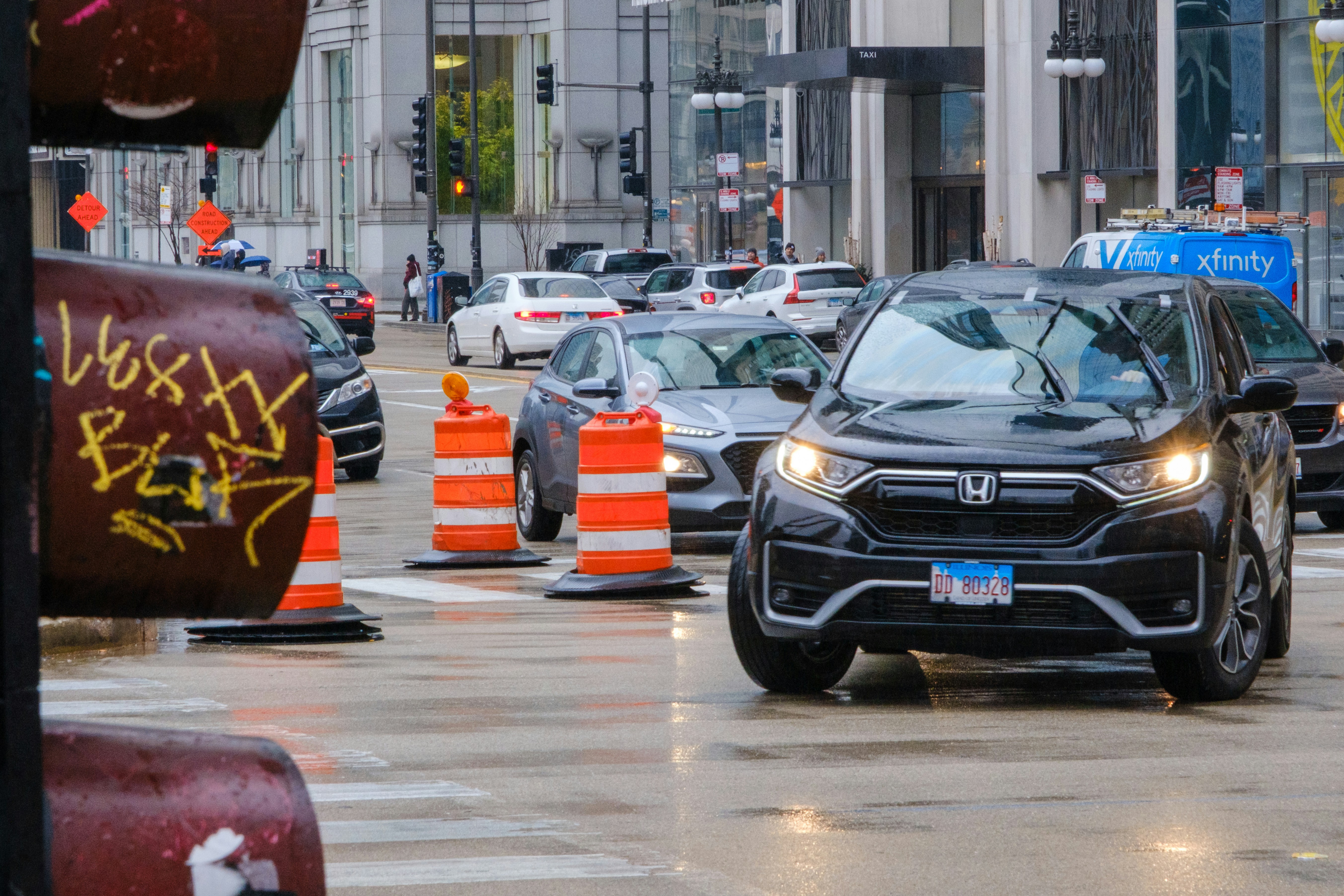 Steel-gray SUV drives through a bustling construction zone on a city street, orange barrels and traffic cones framing the path ahead. Urban storefronts and passing vehicles provide context and motion.
