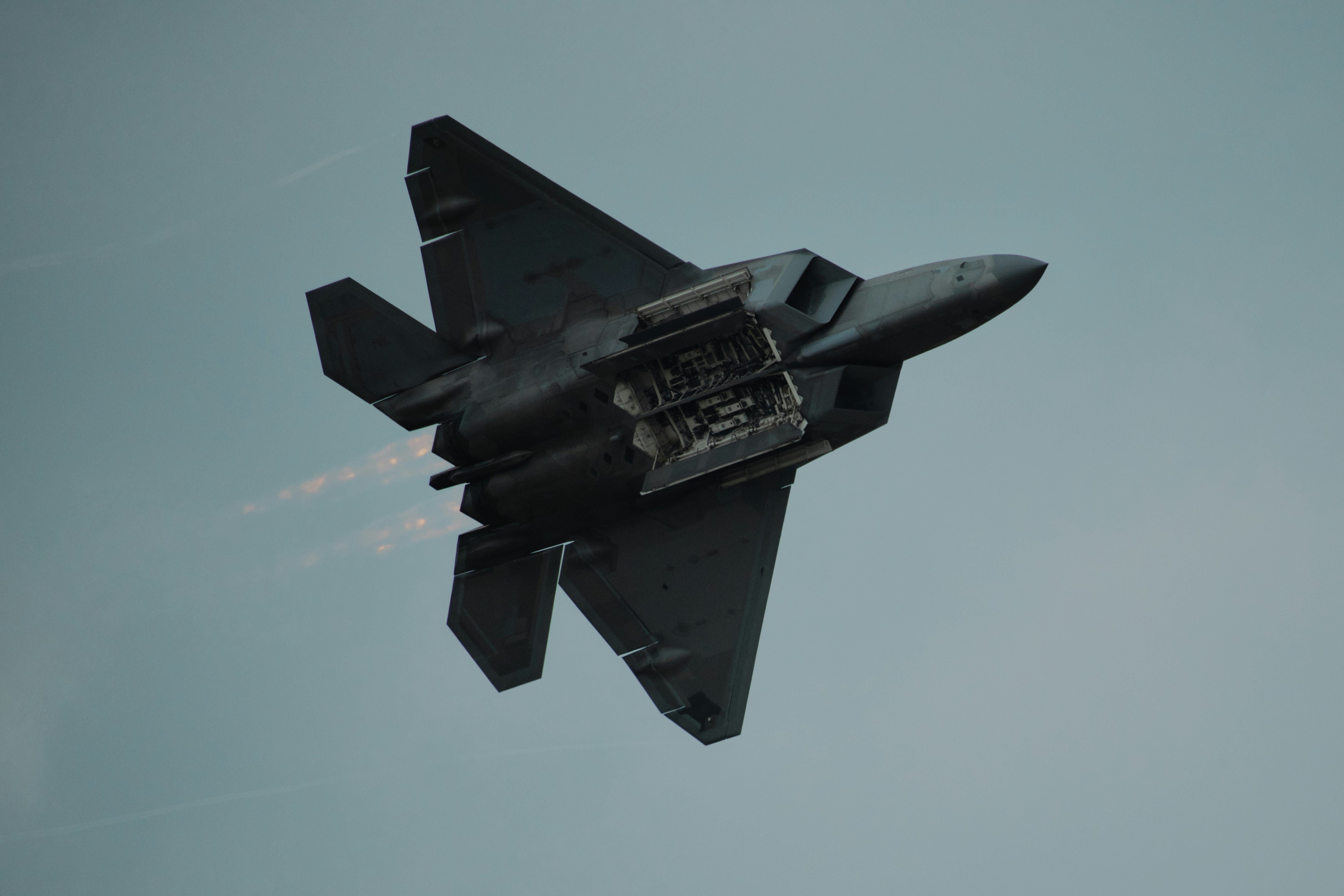 A sleek fighter jet climbs through a clear blue sky, with visible afterburner flames and open landing gear bay details.