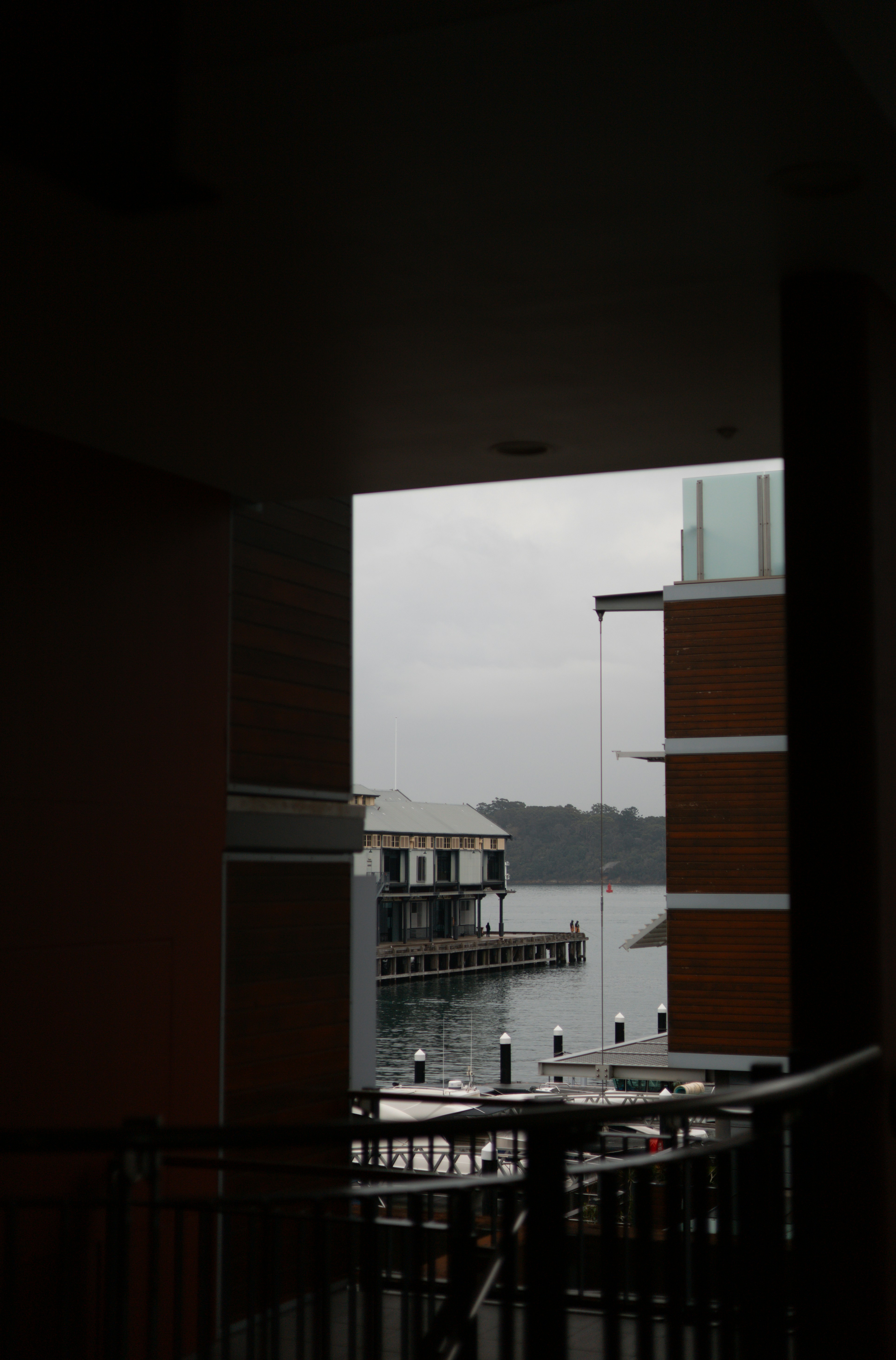 A view of a marina framed by a dark indoor passage, with waterfront buildings and a pier visible through a rectangular opening; the scene includes calm water and a cloudy sky.