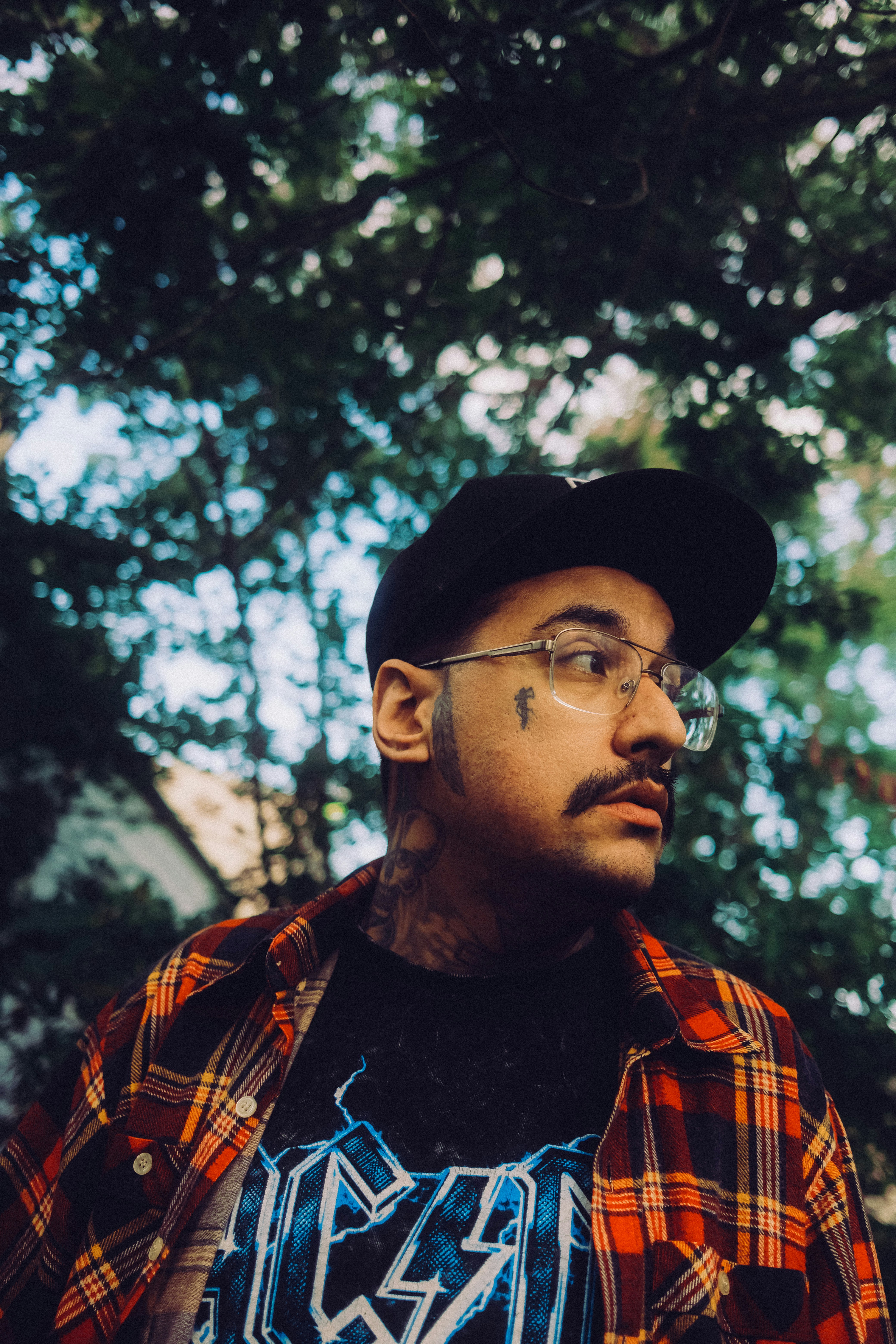 Portrait under Dappled Canopy: A Tattooed Gaze in Plaid and Glass