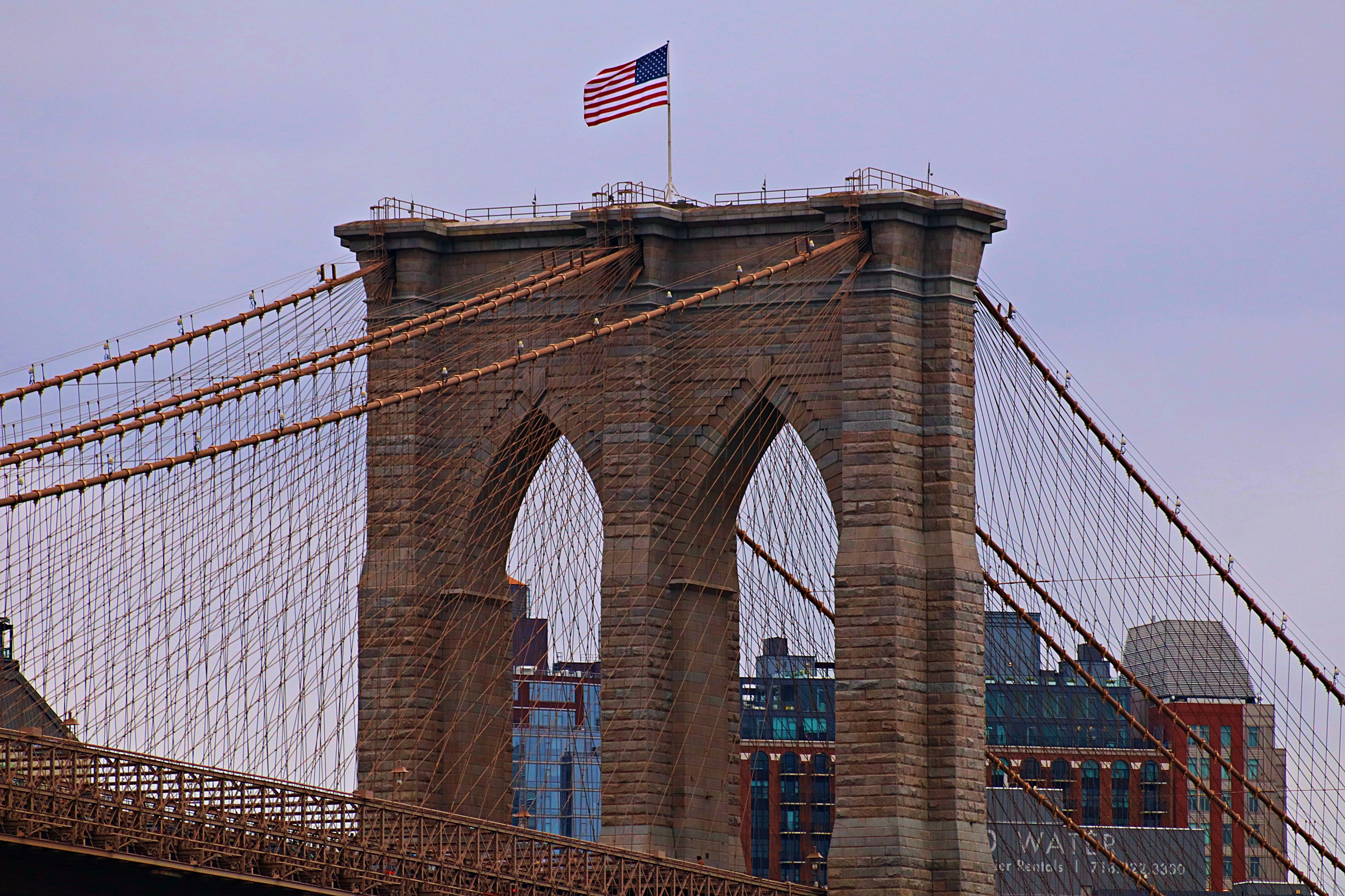 Close-up of the Brooklyn Bridge tower with an American flag, cables fanning out against a pale sky; urban buildings peek behind, conveying a snapshot of city structure and heritage.