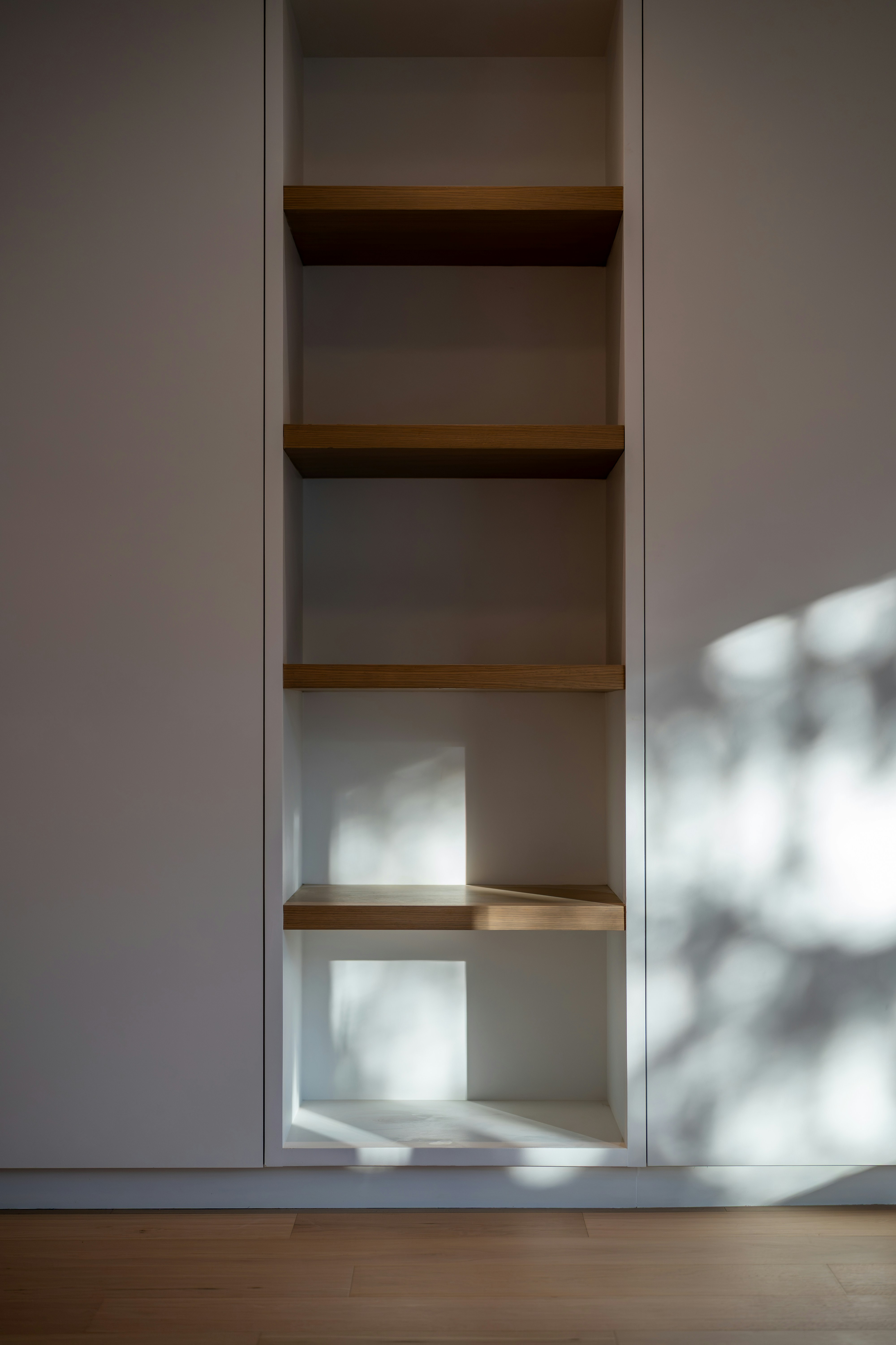 Tall built-in cabinet with wooden shelves, bathed in soft natural light casting geometric shadows along the interior. Subtle reflection on the hardwood floor emphasizes quiet, minimalist space.