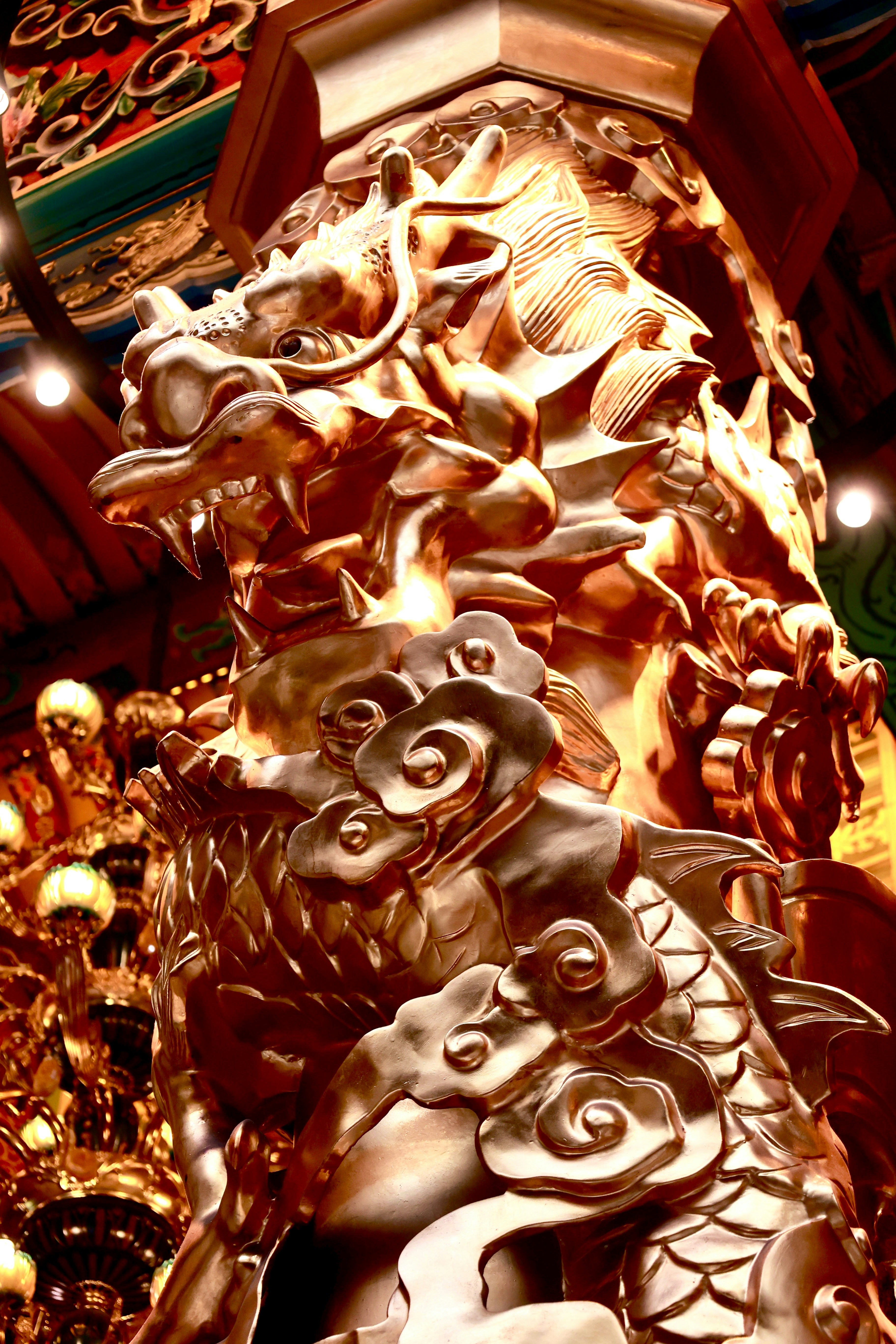 Bronze dragon statue illuminated in a richly decorated interior, showing intricate scales and curling tendrils; the sculpture stands as a centerpiece among ornate decor and warm lighting.