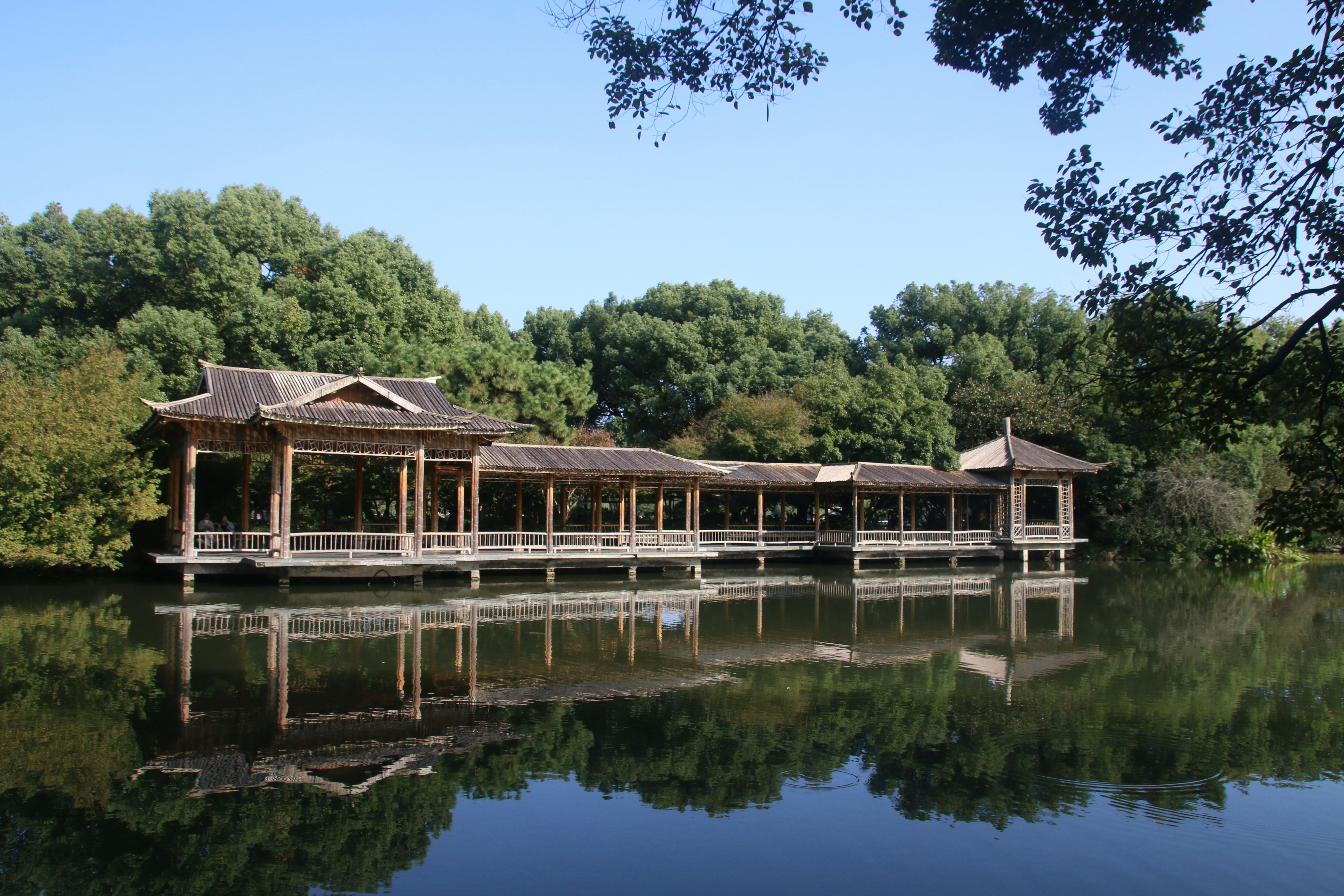 Wooden riverside pavilion stretches across calm lake with tree-lined shore, its reflection crisp on the water’s surface. A serene landscape study emphasizing symmetry and natural light. Note: photograph captures a traditional structure and its mirror image.