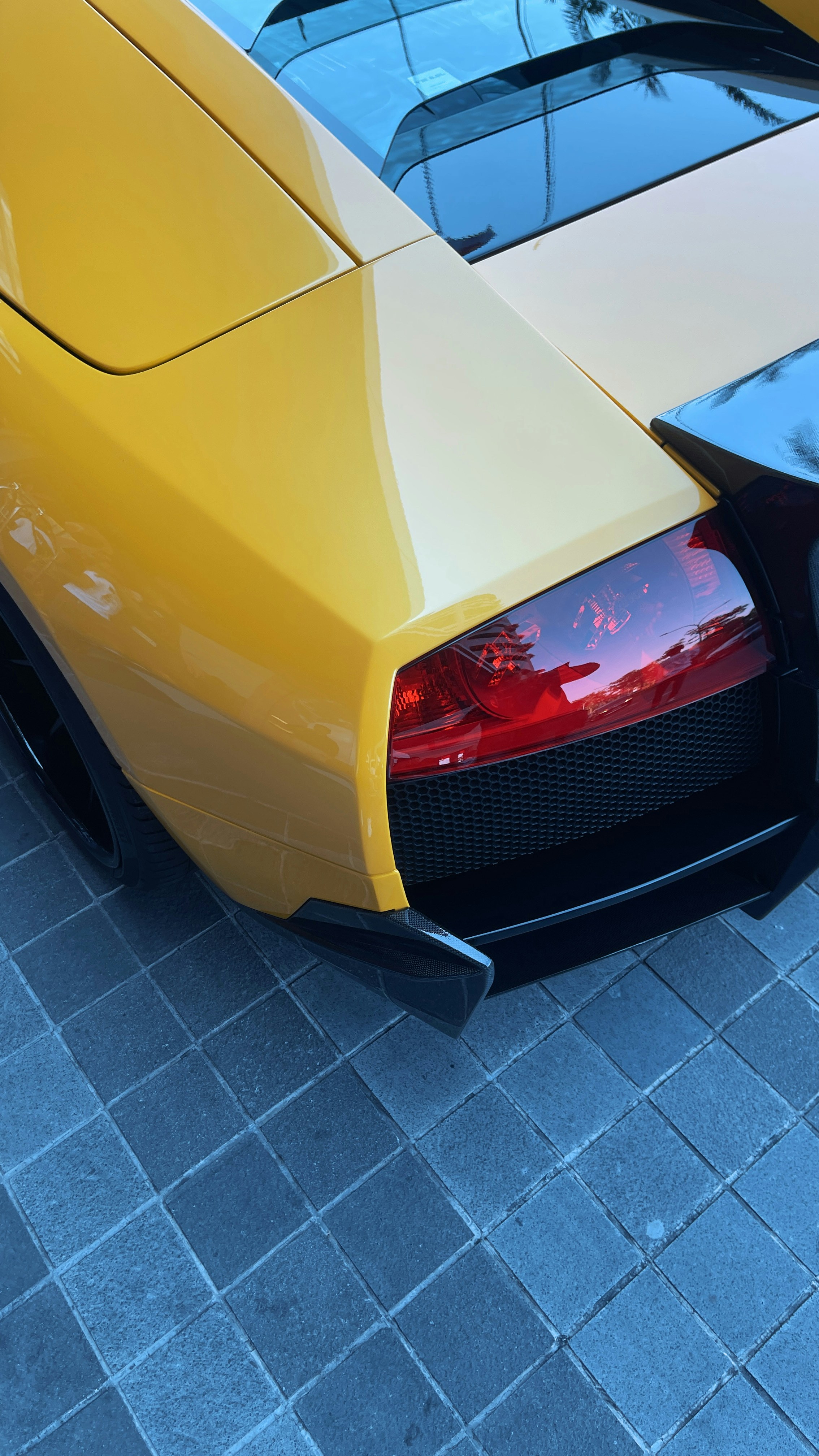 Vibrant yellow rear quarter of a low-slung sportscar with red taillight and exposed aero details; captured on a tiled surface, focusing on the body lines and ventilation mesh.