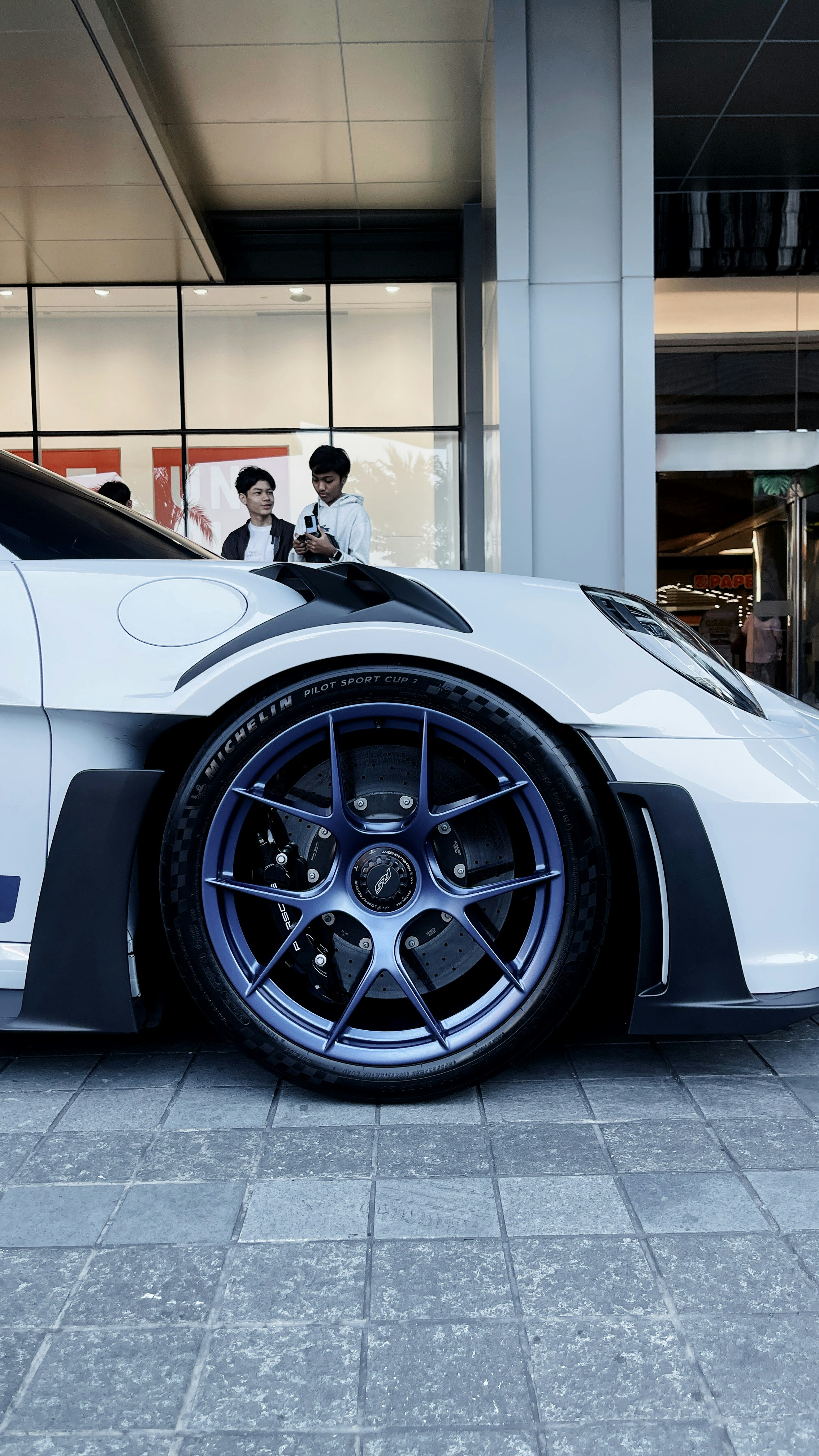 Low-angle view of a white sports car with blue rims beside a modern storefront